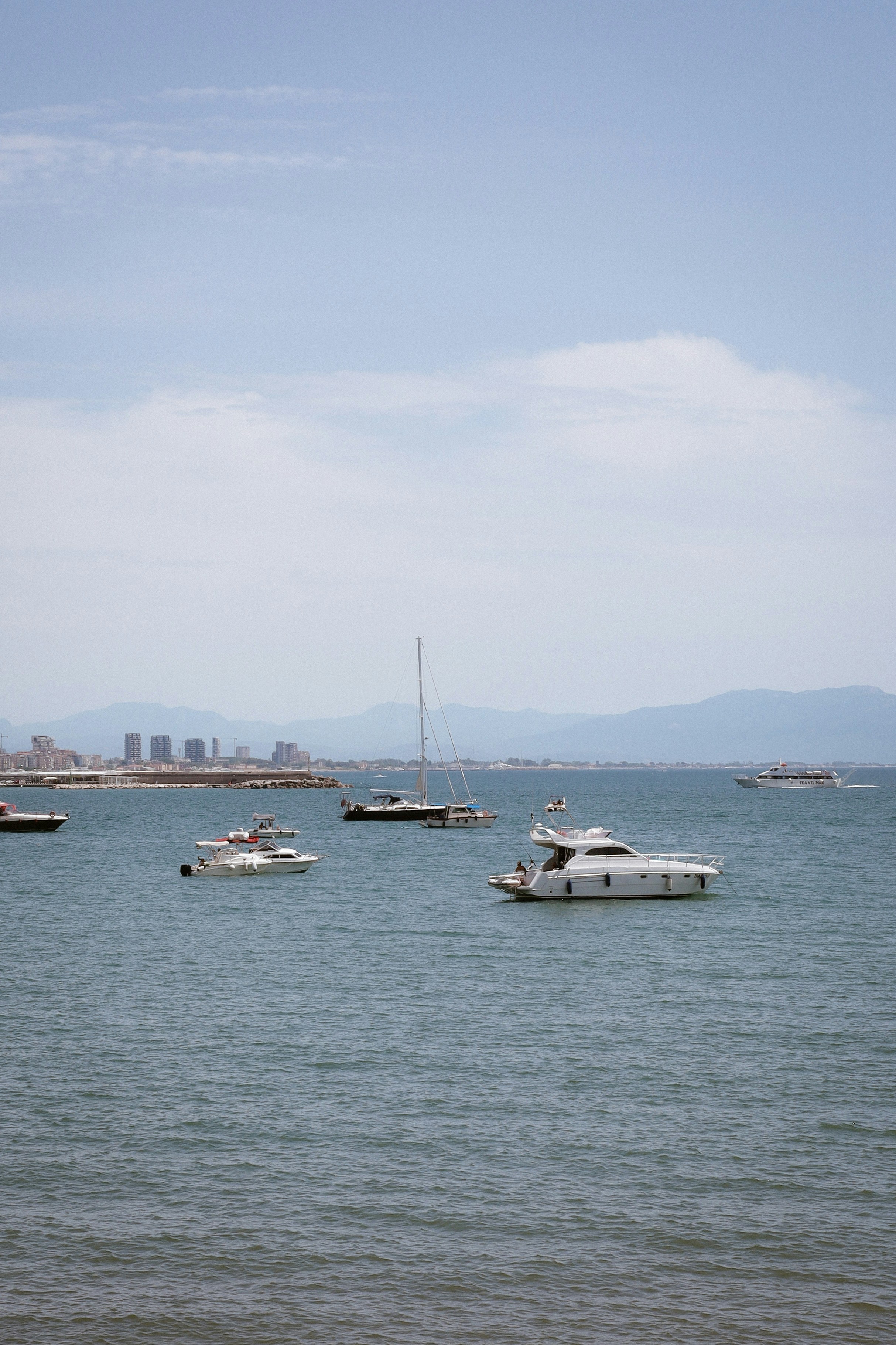 A serene harbor scene featuring various boats gently floating on calm waters, with a distant city skyline and mountains in the background.