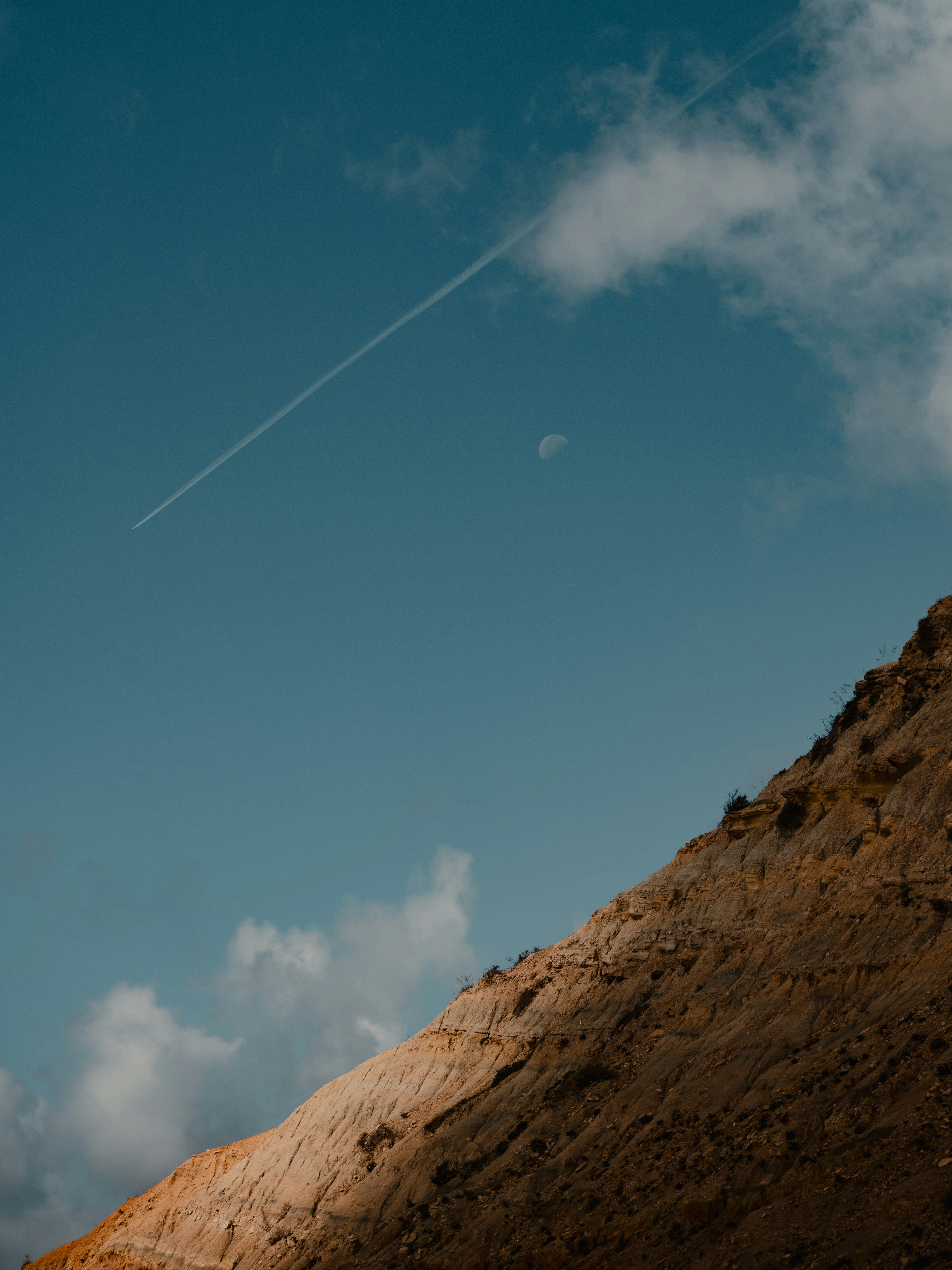 A rugged mountain slope bathed in warm light contrasts against a deep blue sky, where a faint crescent moon and a jet trail add intrigue.