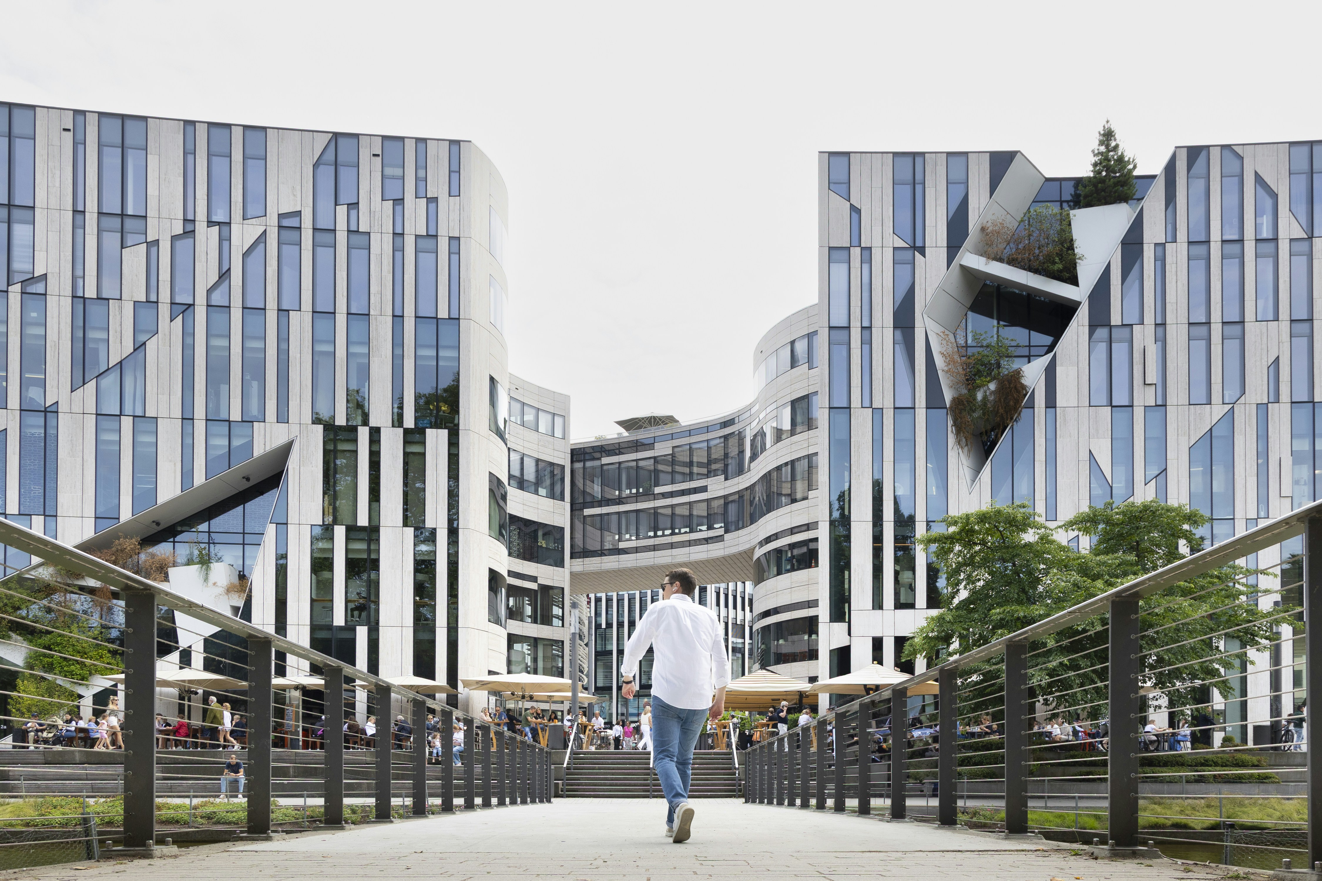 A modern pedestrian walks toward the architectural landmark Kö-Bogen shopping complex in Düsseldorf, Germany — a hub of design, shopping, and urban life. | Person walks towards modern buildings on a bridge.