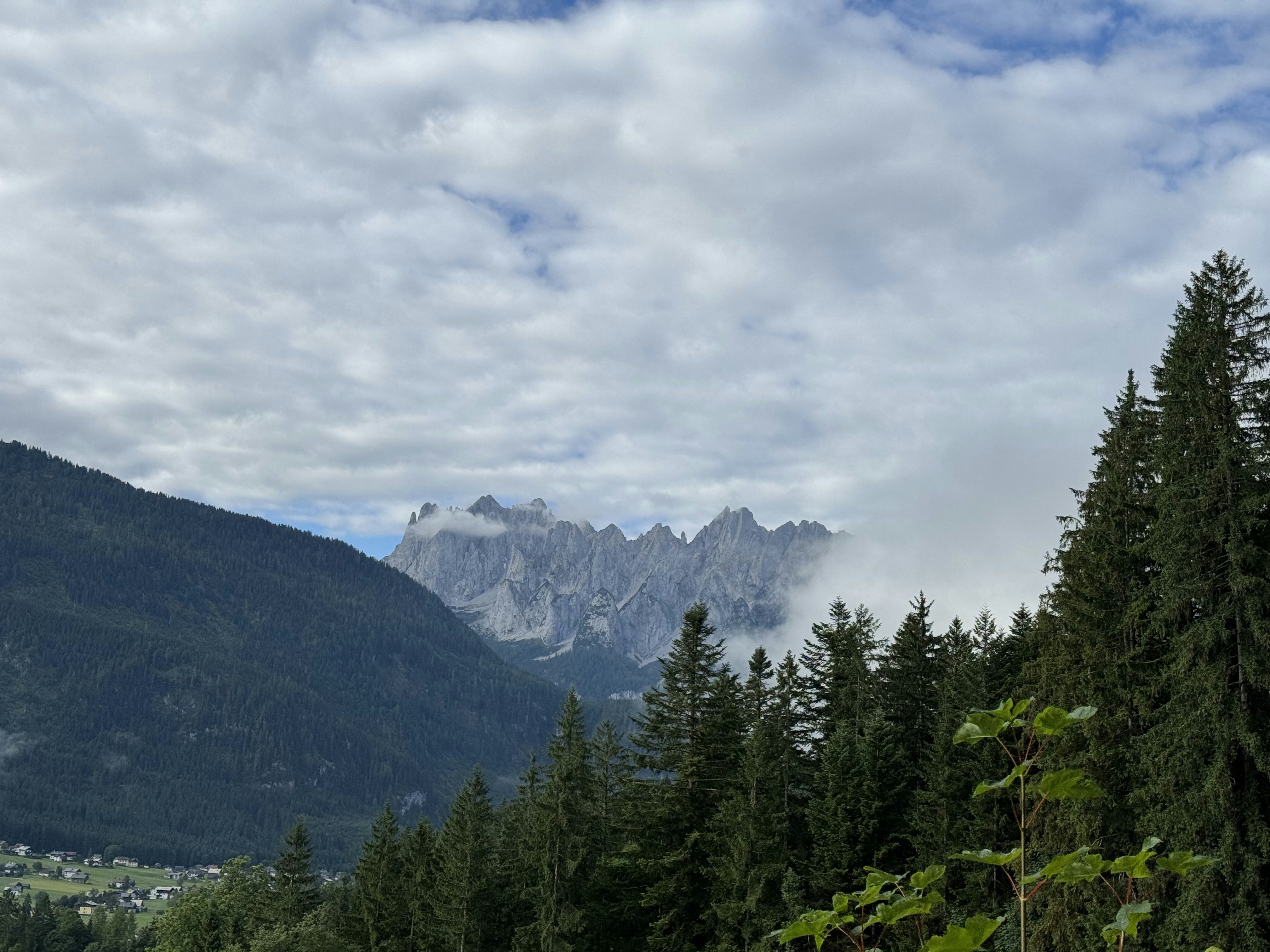 Dramatic mountain range shrouded in clouds, framed by lush evergreen trees and a serene valley below.