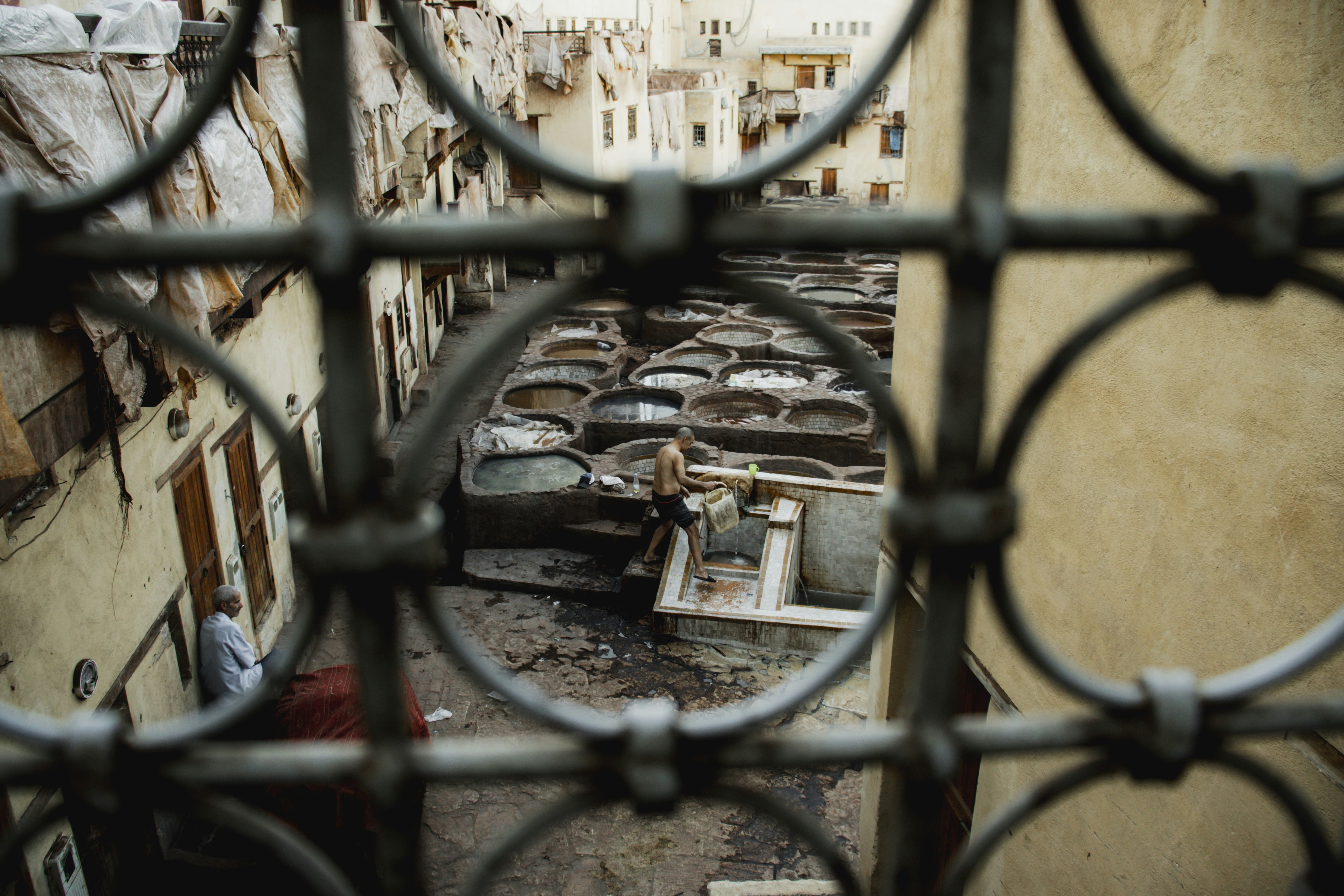 A craftsman working in a historic tannery, surrounded by circular dyeing vats, framed by an ornate metal grille. The scene captures the essence of traditional leather-making techniques.