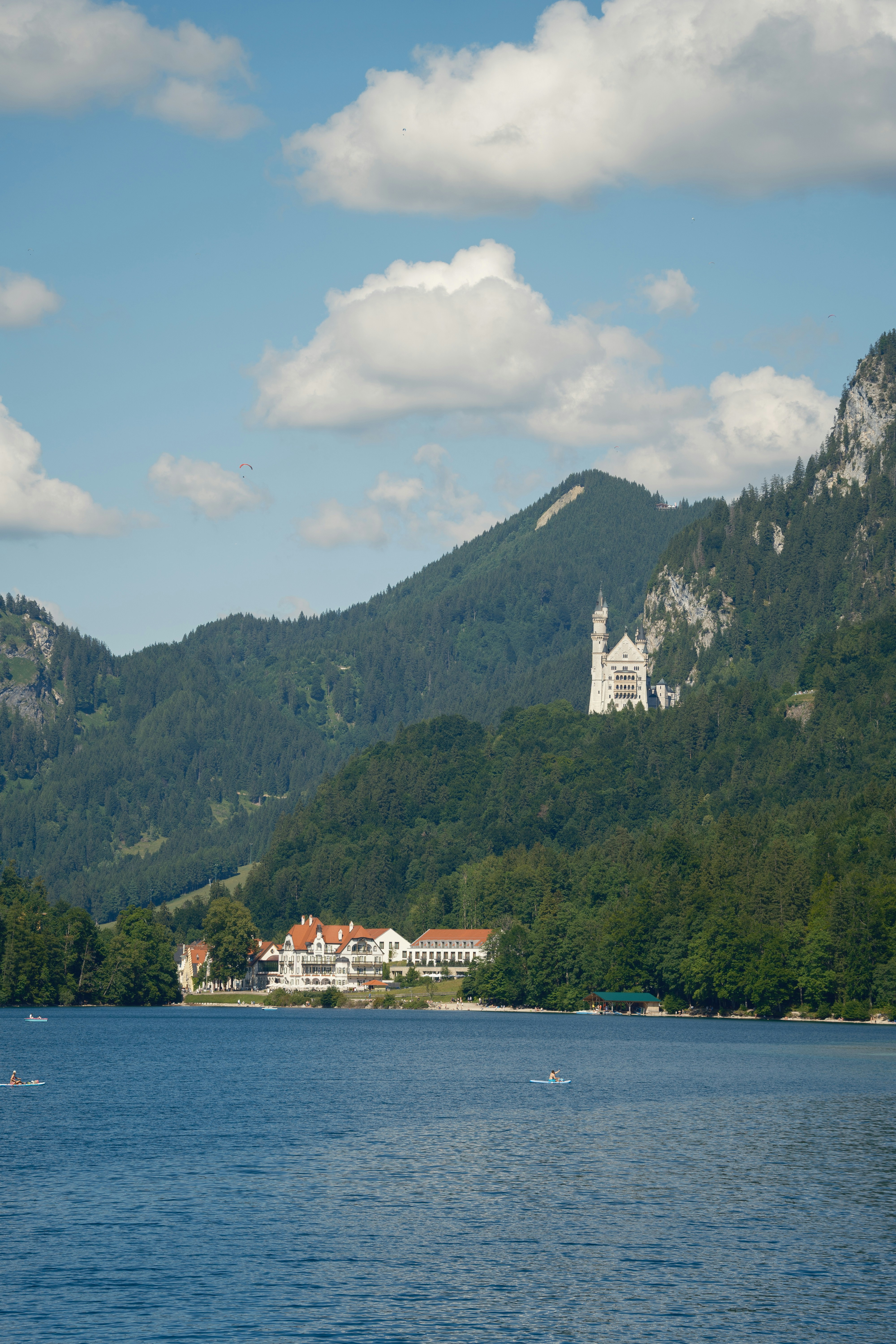 A picturesque view of a grand castle nestled among verdant mountains, overlooking a serene lake with kayakers gliding across the water.