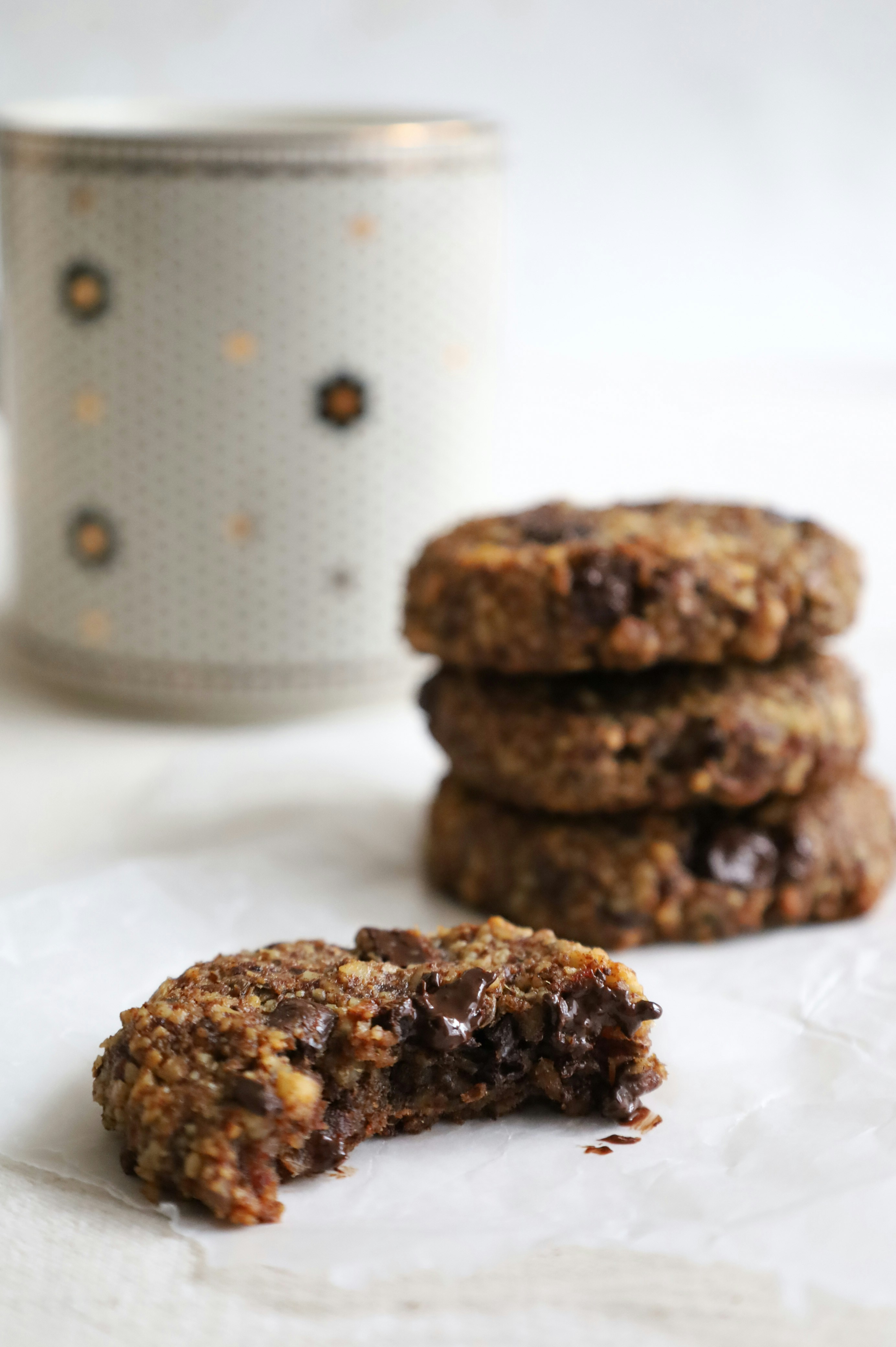 A stack of freshly baked cookies with a bite taken out, revealing gooey chocolate inside, accompanied by an elegant patterned mug in the background.