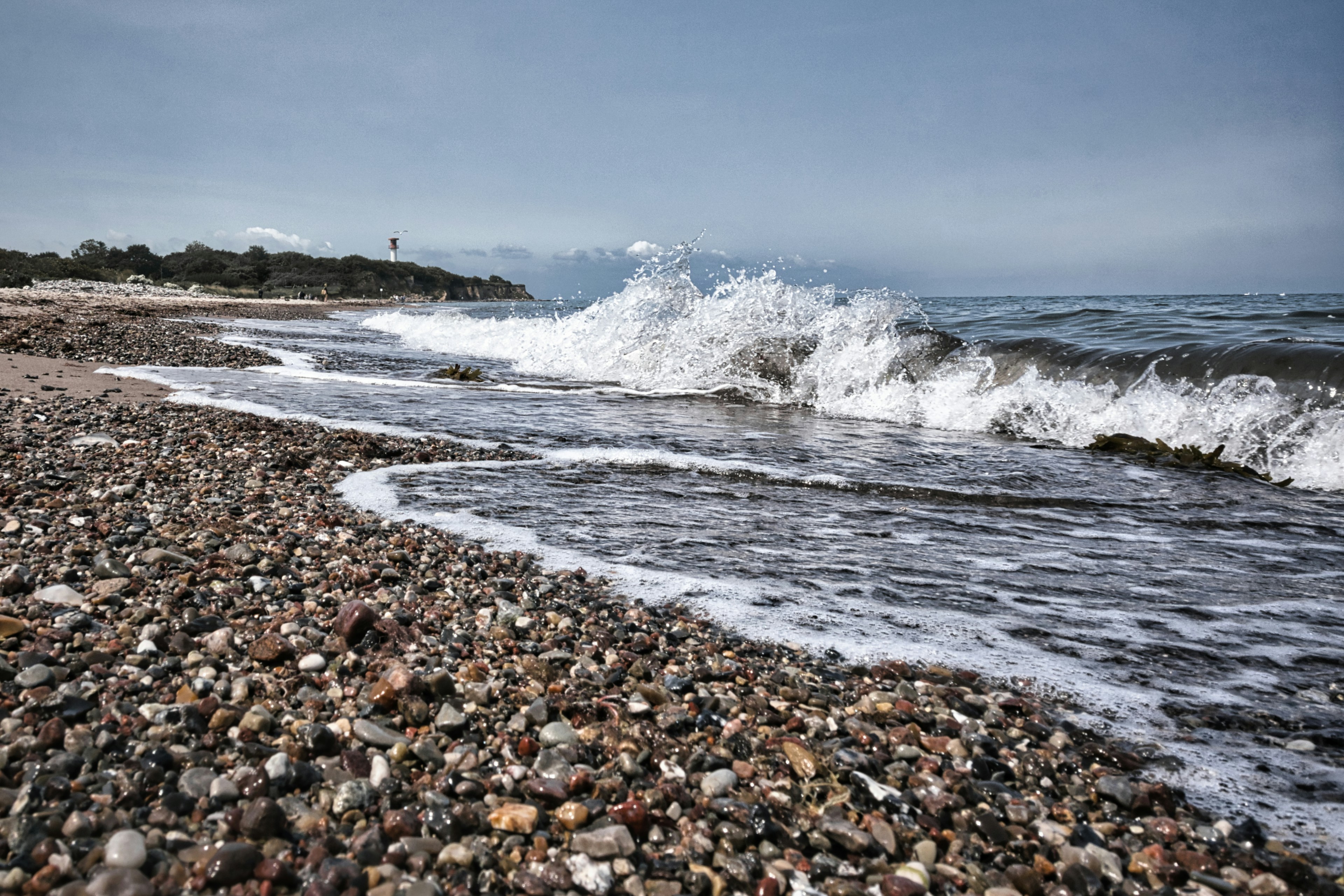 Waves crashing against a pebbled beach under a cloudy sky, with a distant lighthouse visible on the horizon.