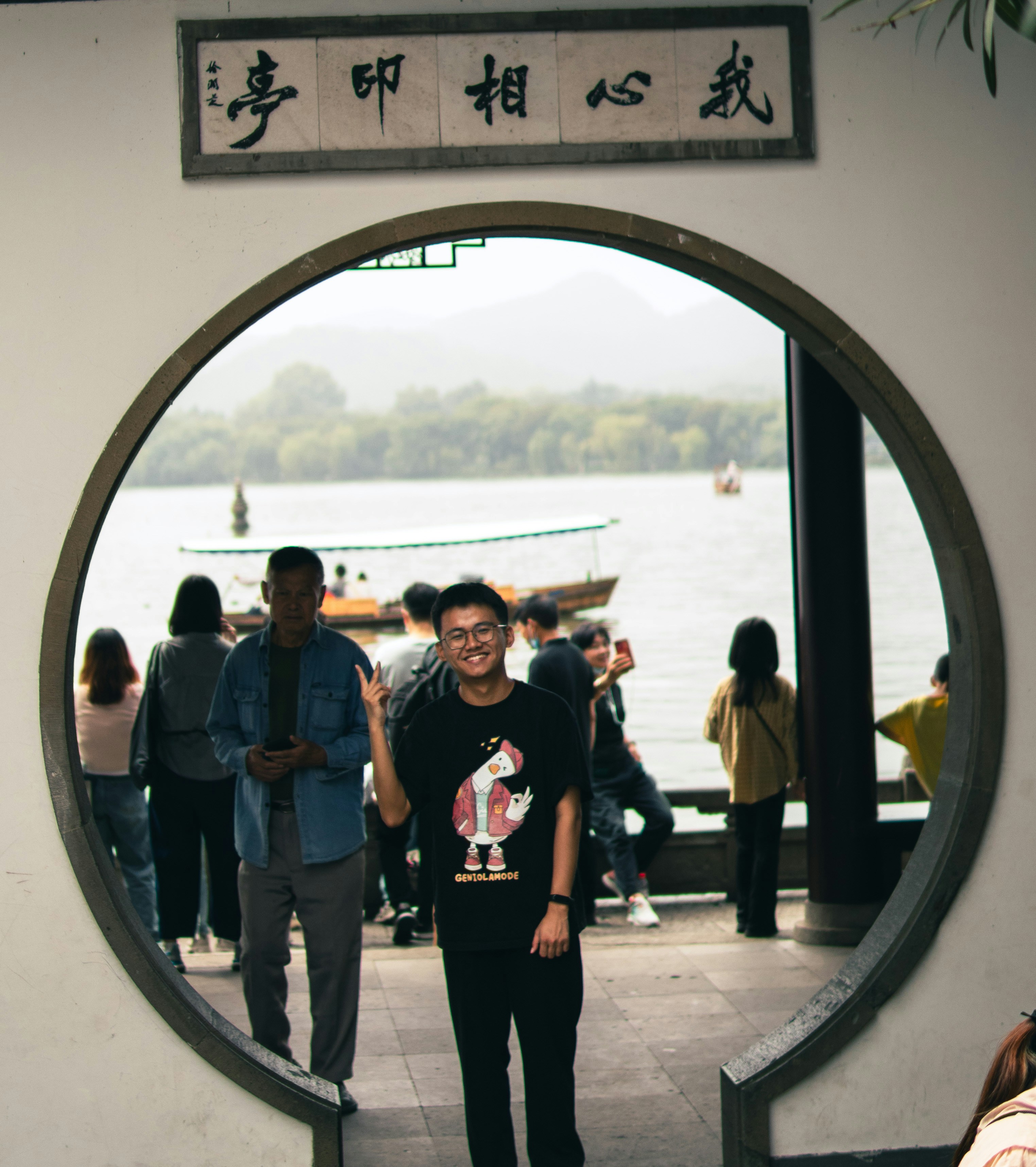 A cheerful young man poses in a circular archway, framed by visitors and a serene lake view in the background. The scene captures a moment of connection and leisure.