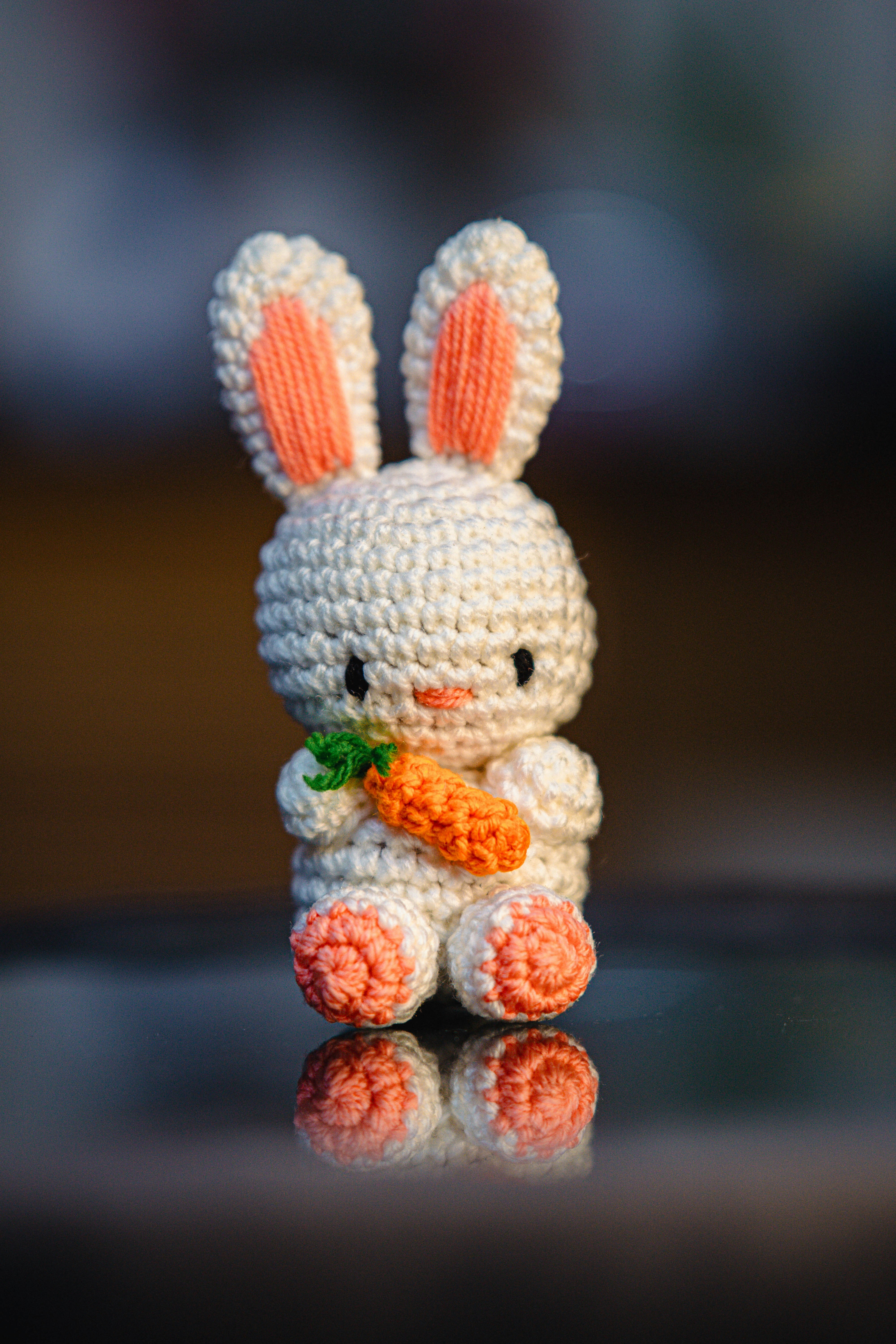 A cute crochet bunny posed alongside bright yellow sunflowers, captured in soft natural light. This handmade scene blends warmth, charm, and artisan craftsmanship — ideal for themes like nursery decor, eco-friendly toys, or whimsical product photography. | Here is a caption for the image: a crochet bunny holding a tiny carrot.