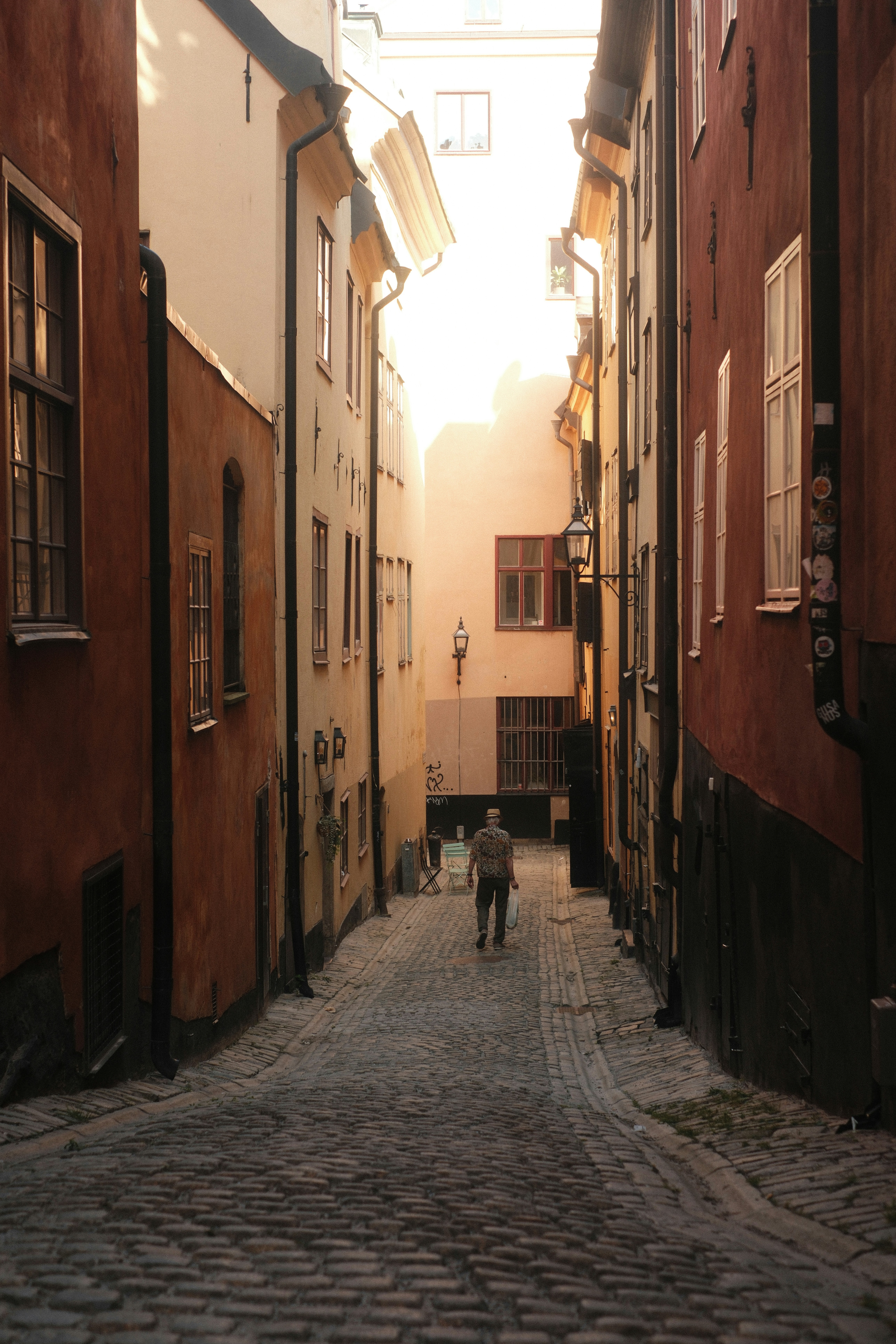 A solitary figure walks down a narrow cobblestone street flanked by warm-toned buildings, capturing the charm of an old town. The sunlight filters softly through the alley, enhancing the tranquil atmosphere.