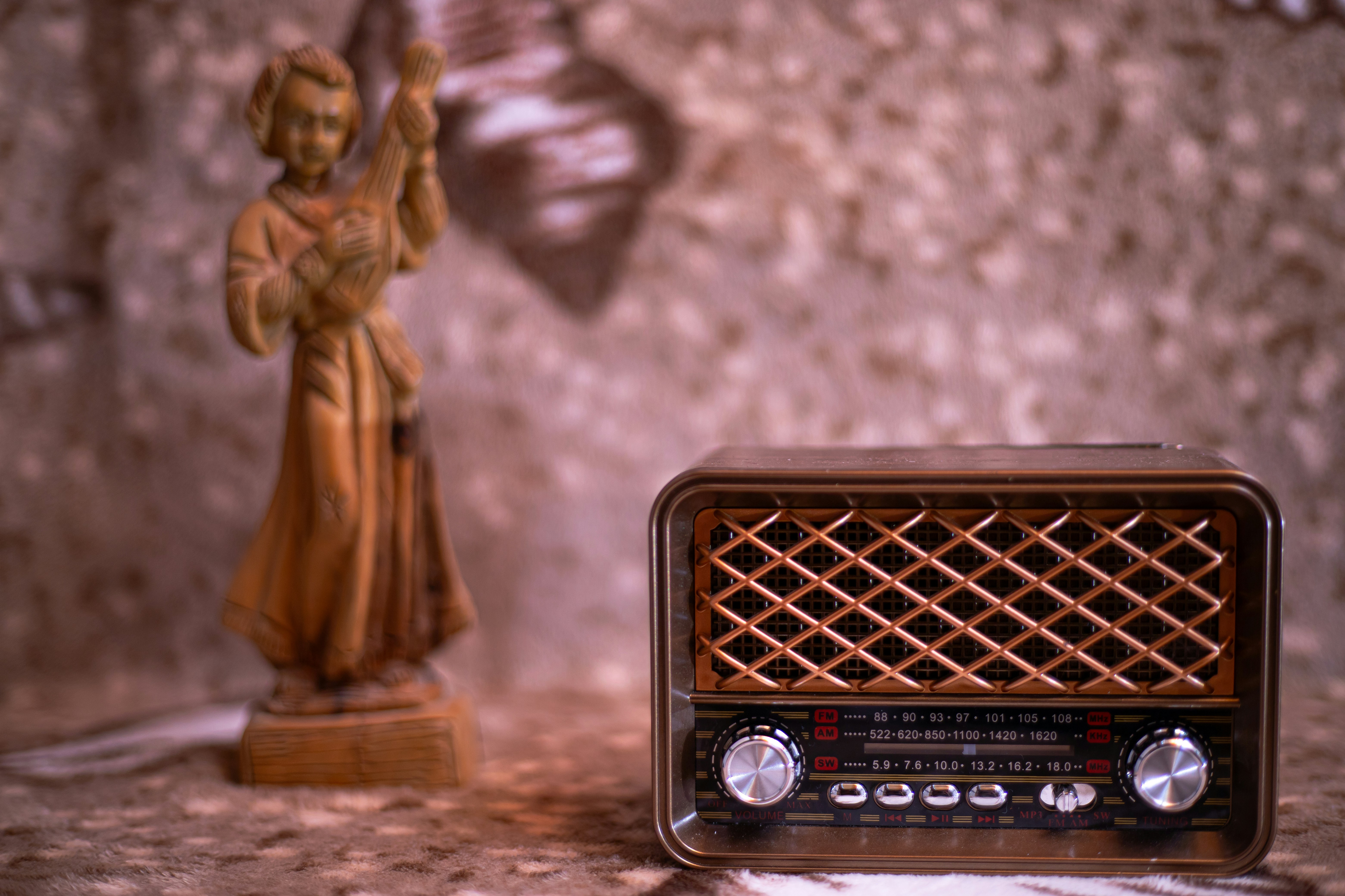 A vintage-style radio with a brown lattice grille and analog dials sits beside a carved wooden statue of a musician holding a stringed instrument. The scene blends retro technology with handcrafted artistry, evoking a nostalgic atmosphere where sound and sculpture meet. | A vintage radio sits next to a wooden figurine.