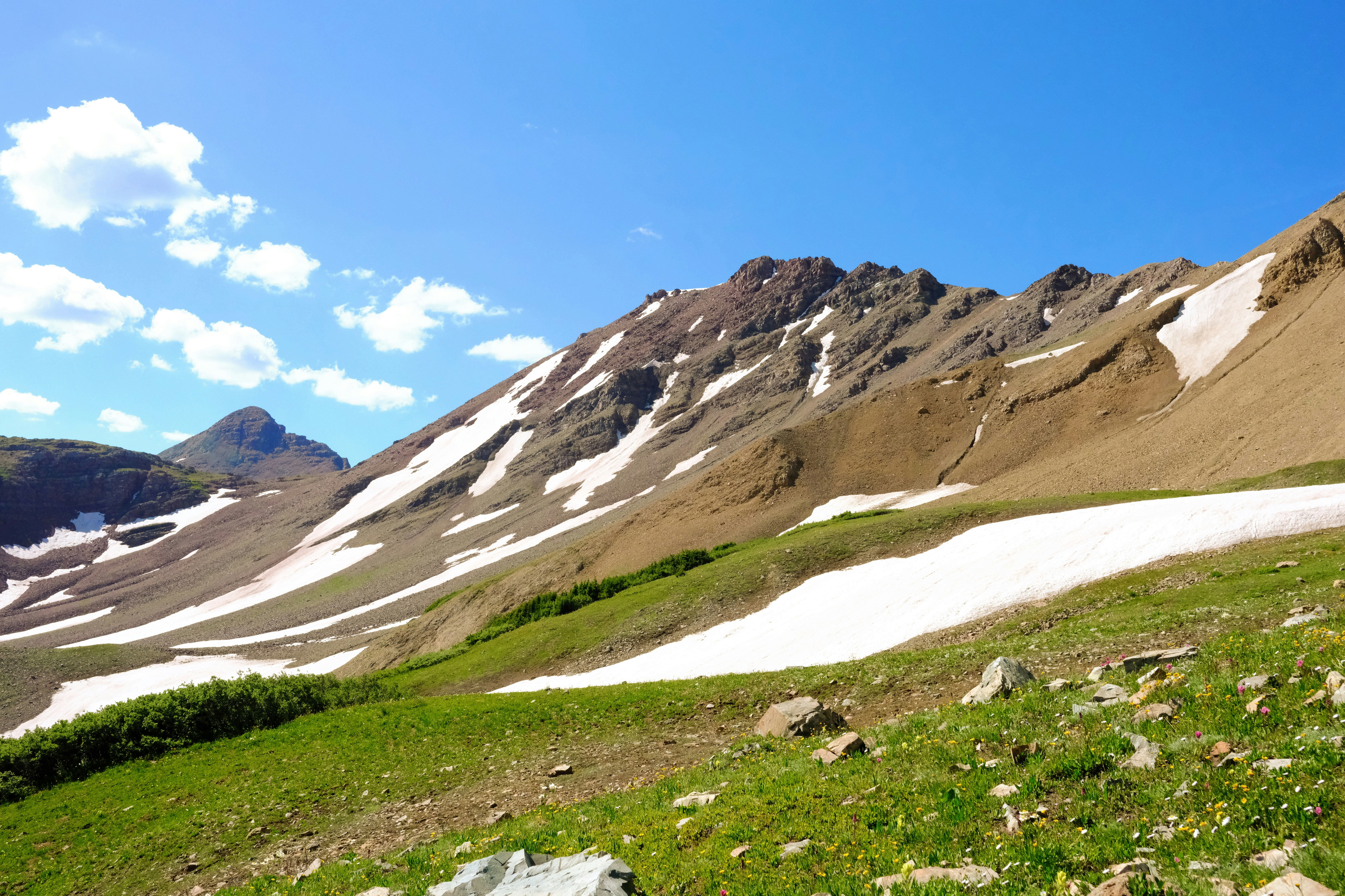 Vast mountain terrain showcasing patches of snow and verdant grass under a bright blue sky.