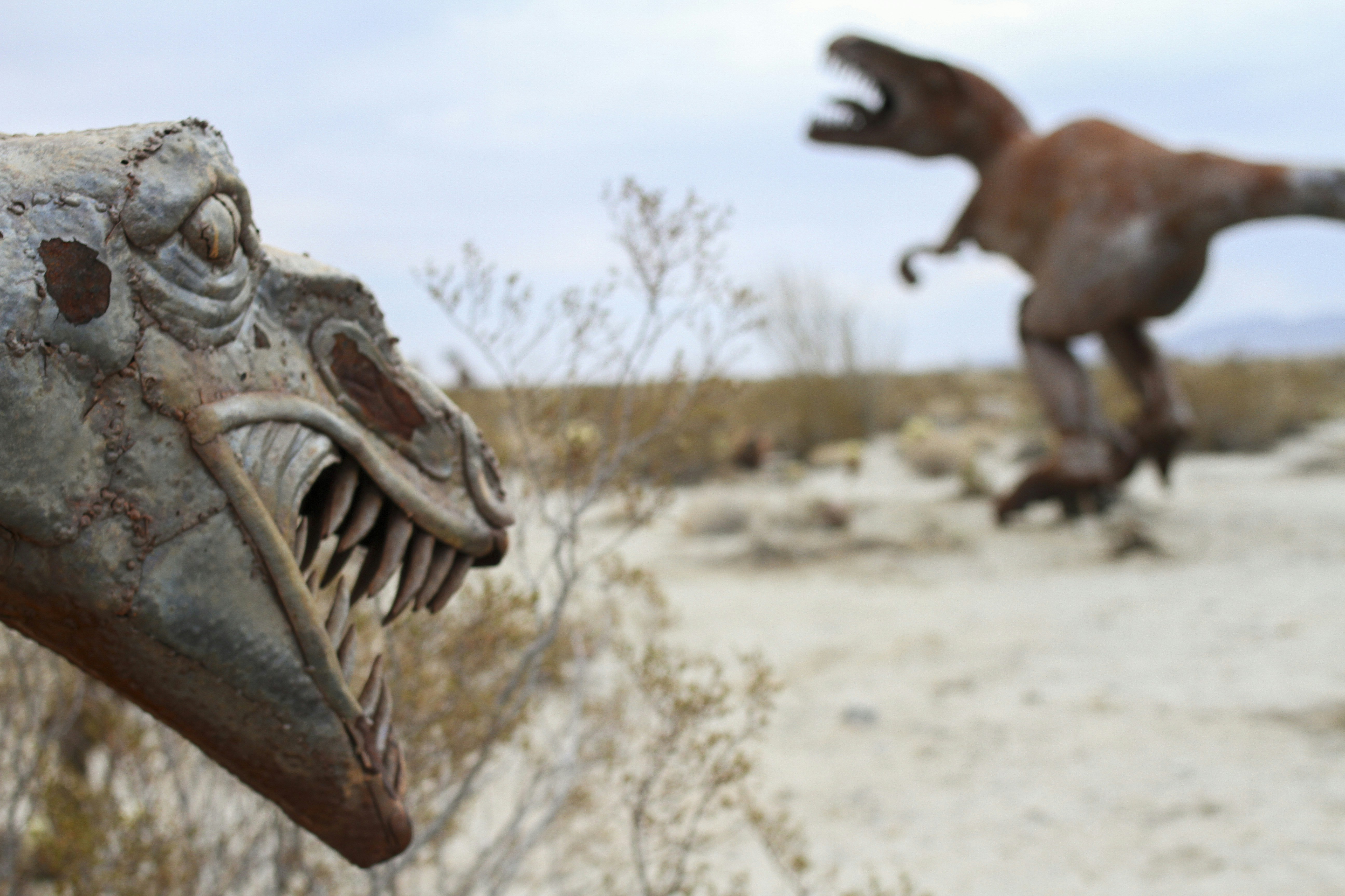 A detailed dinosaur sculpture in the foreground, with a larger, dynamic dinosaur figure in the background, set against a barren desert landscape.