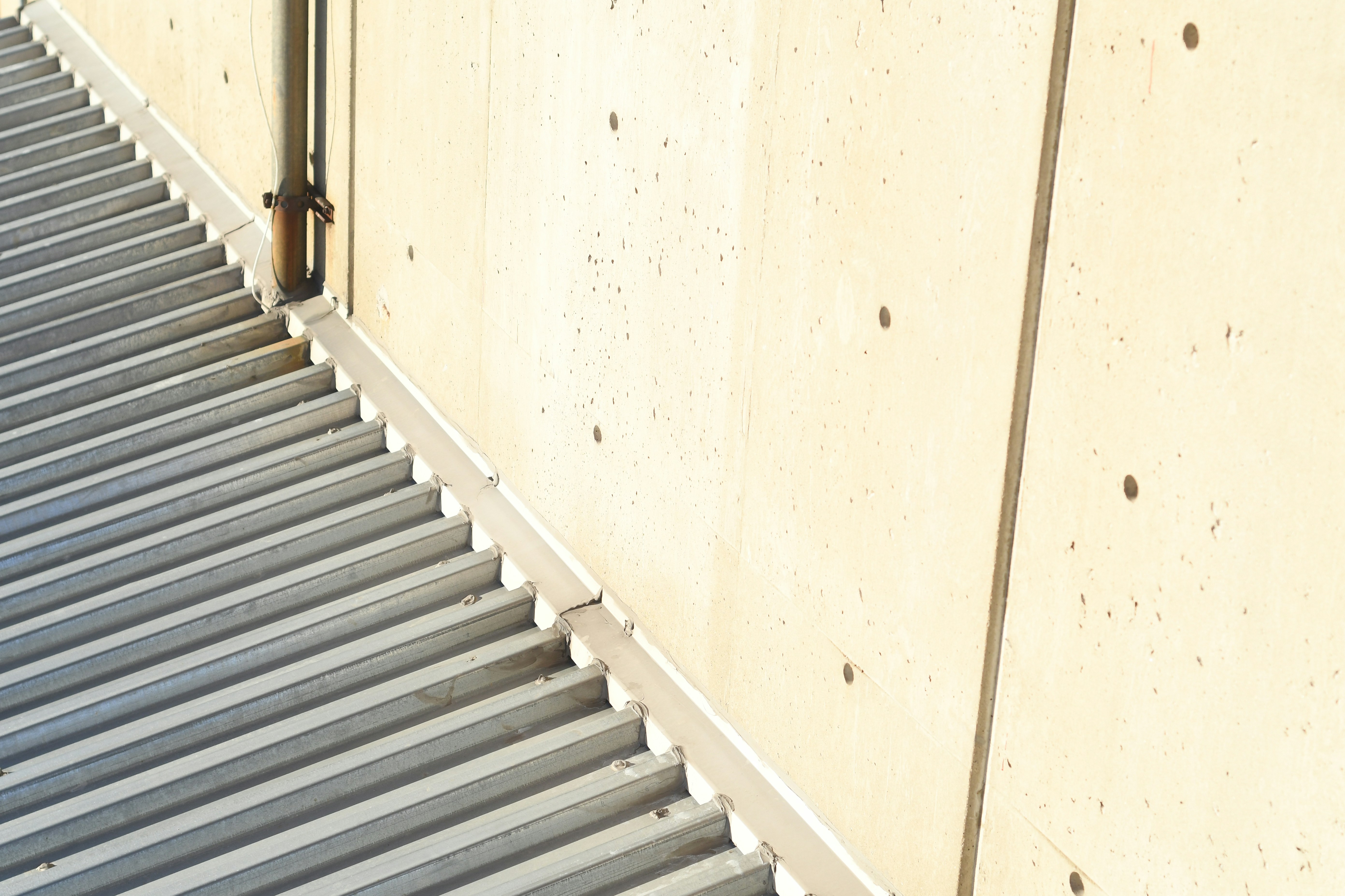 Metal roofing panels converge with a textured concrete wall, showcasing a blend of industrial materials. The play of light emphasizes the structural details.