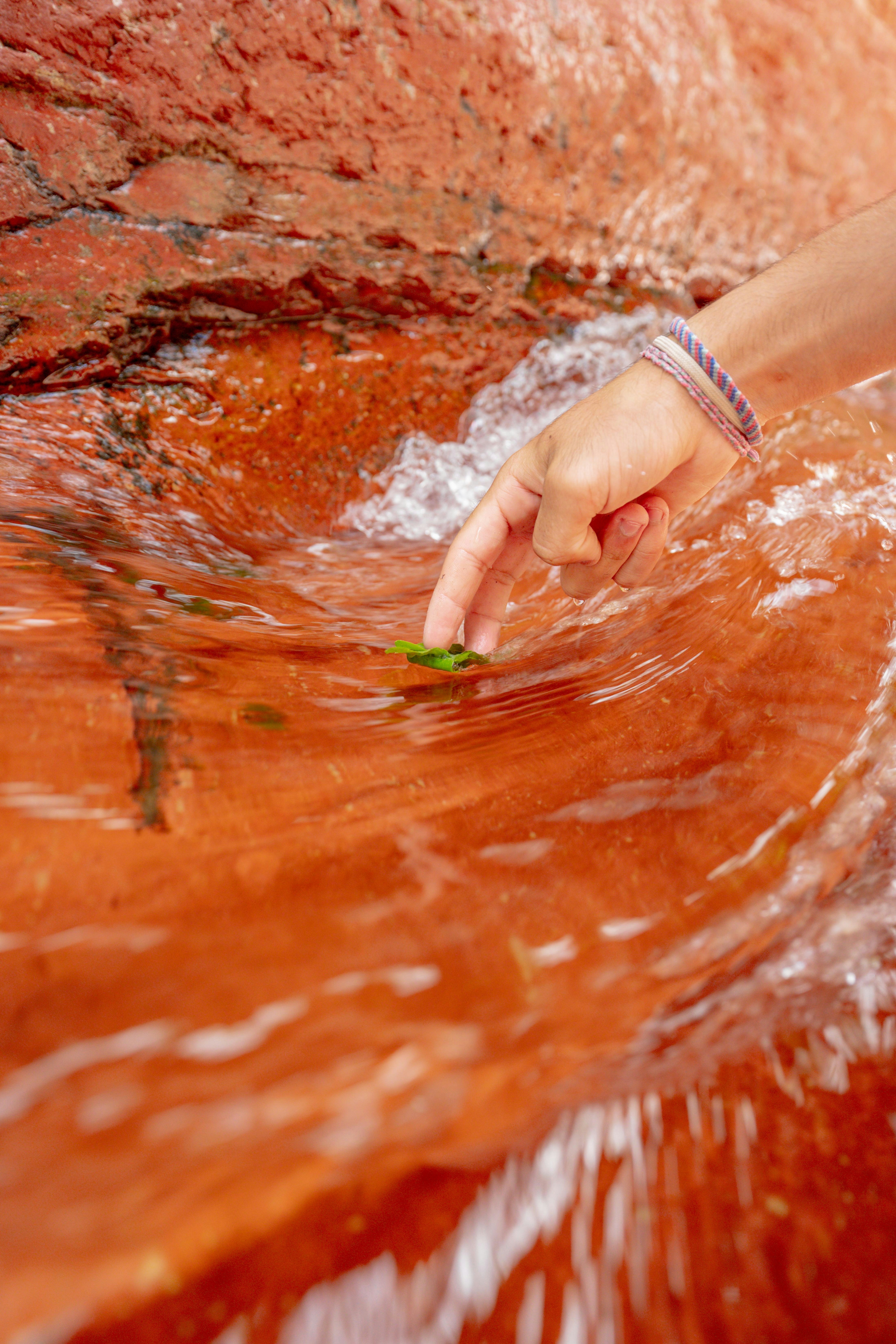 A hand delicately reaches into a flowing stream, touching a vibrant green leaf amidst the rippling water. The warm tones of the surrounding rocks enhance the serene atmosphere.