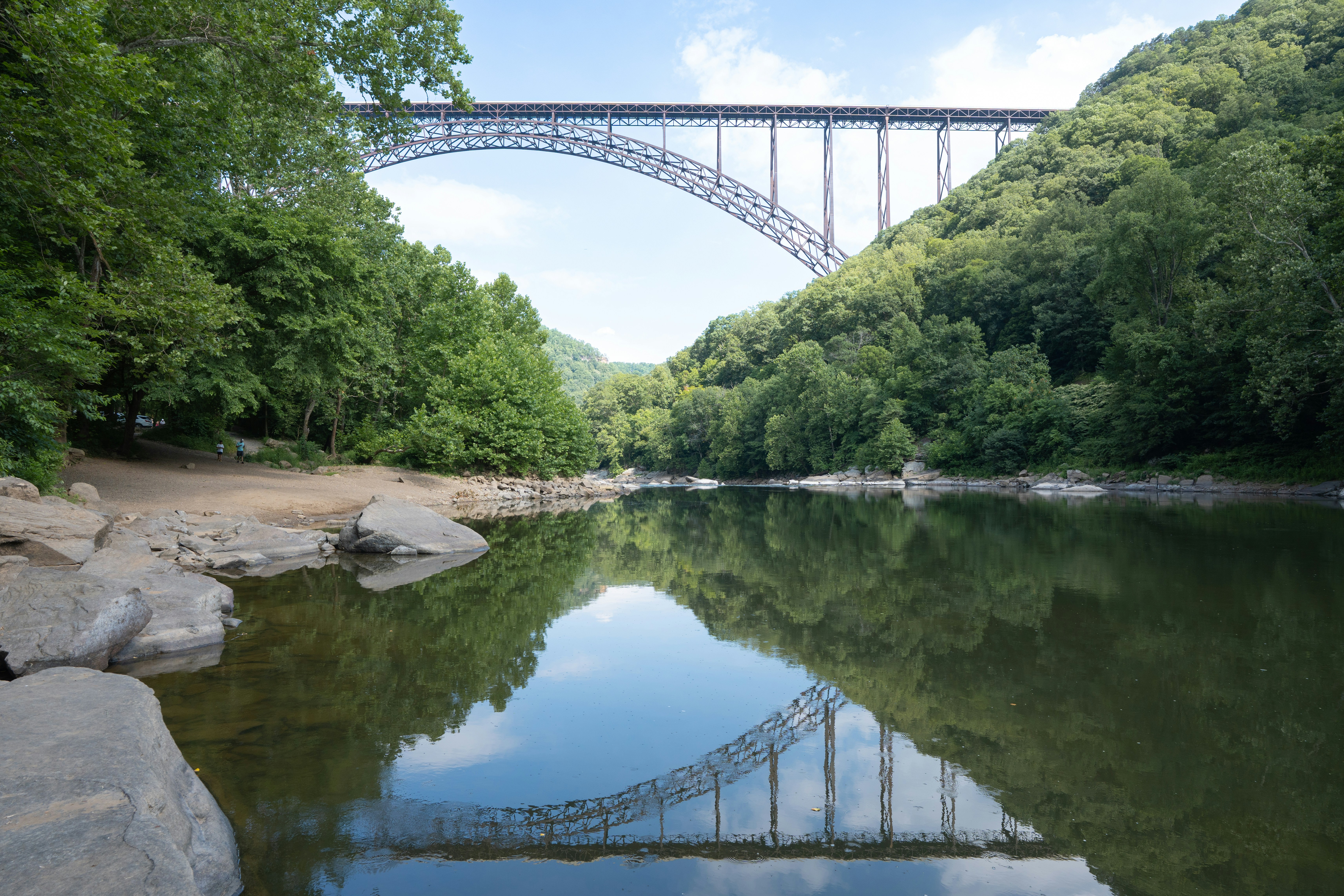 A majestic bridge spans a serene river, mirrored perfectly in the calm waters below, surrounded by lush greenery.