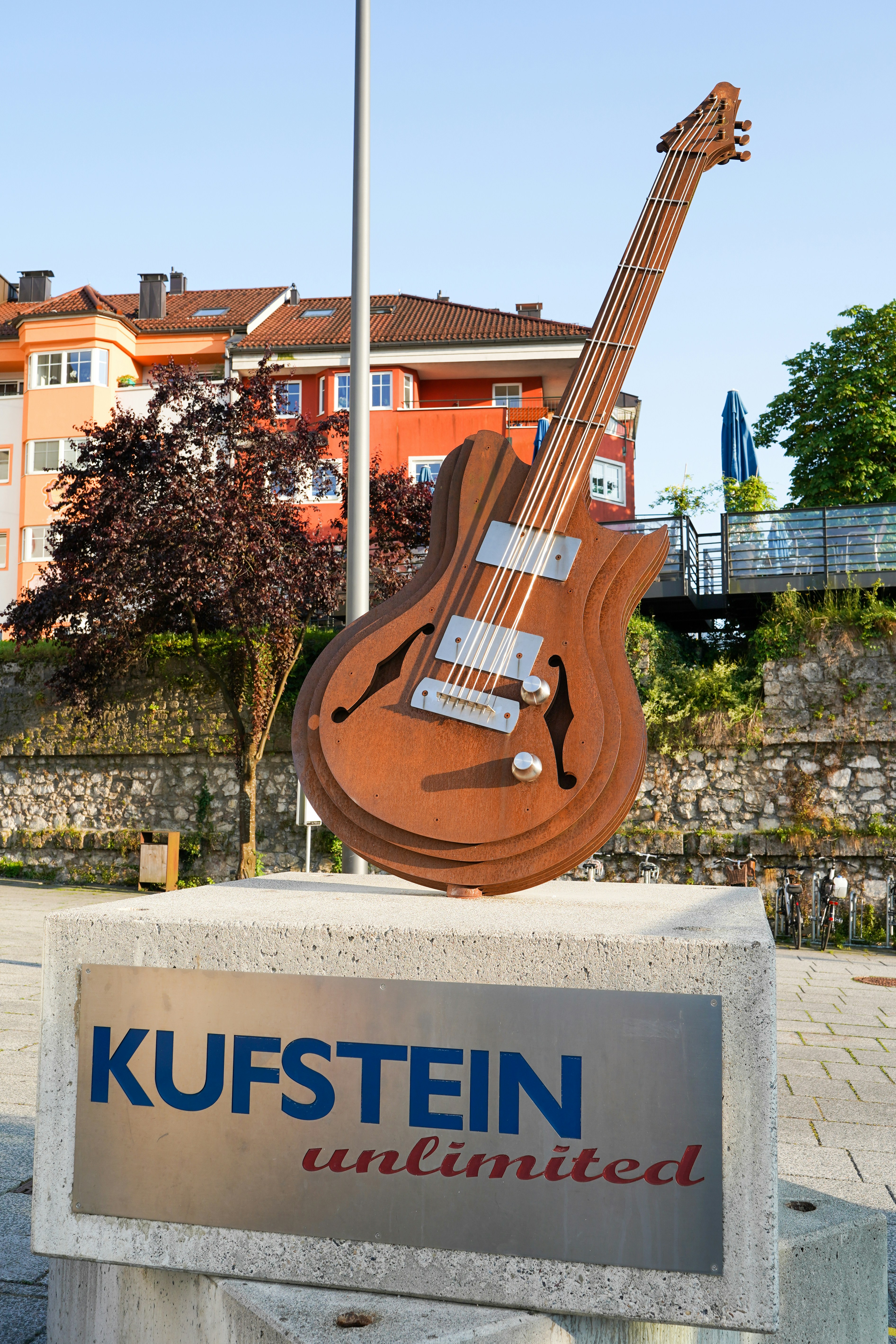A wooden guitar sculpture mounted on a stone pedestal, accompanied by a sign that reads 'KUFSTEIN unlimited.' The backdrop features colorful buildings and greenery.