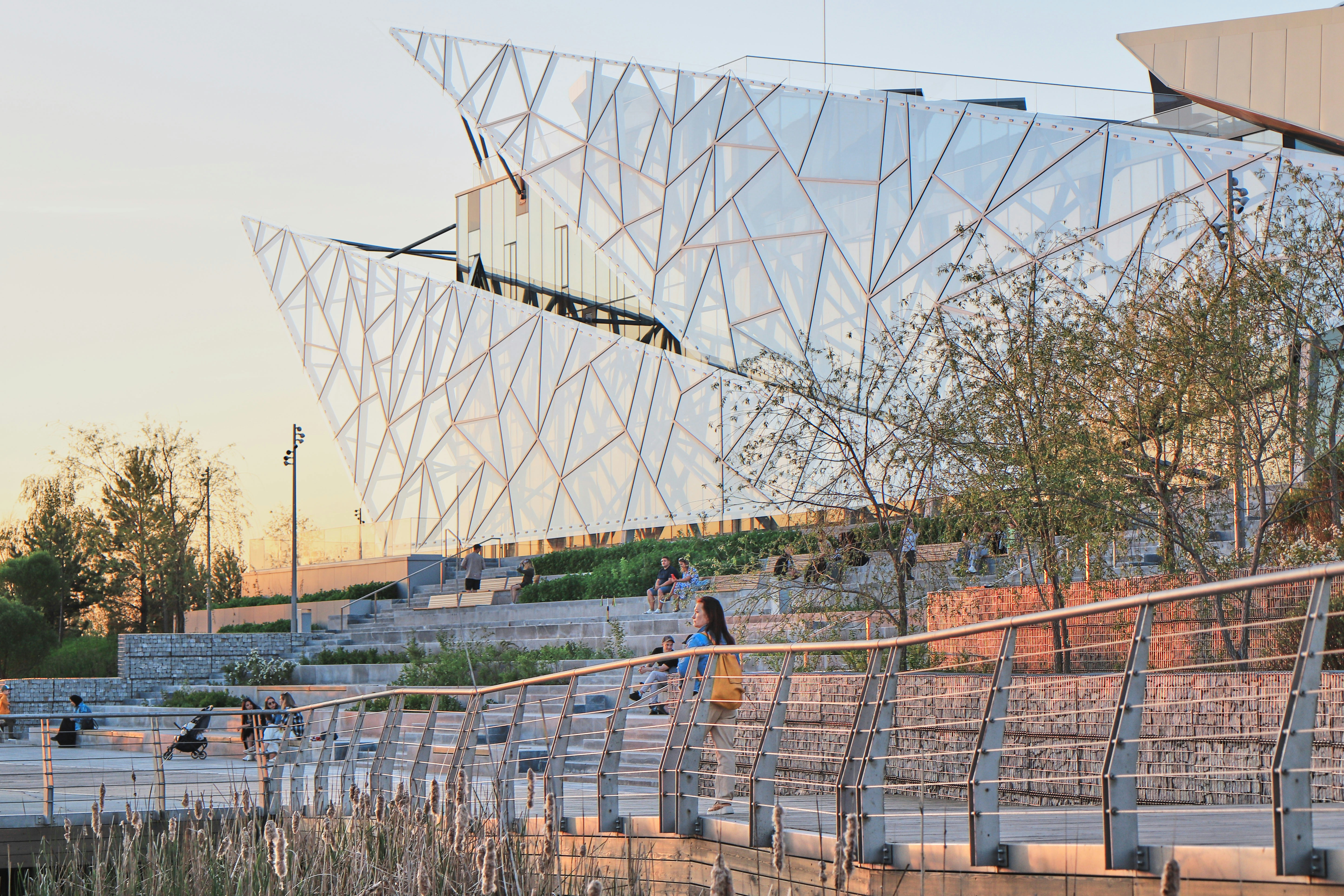 Contemporary building with angular glass facades reflecting warm sunset light, alongside a serene walkway and greenery.