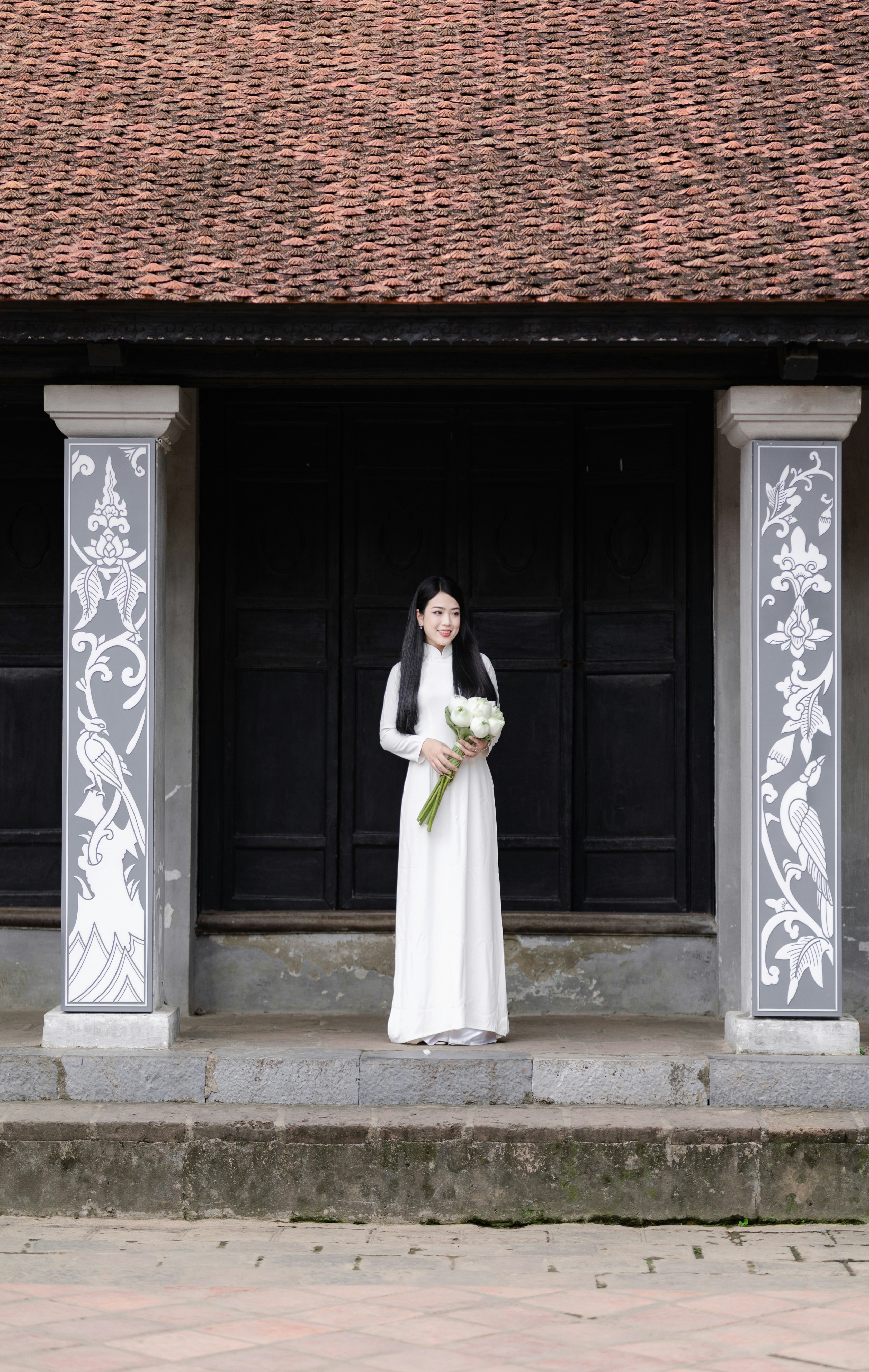 A woman in a traditional white dress stands gracefully with a bouquet in front of an intricately designed doorway. The contrasting textures and colors highlight her poise.