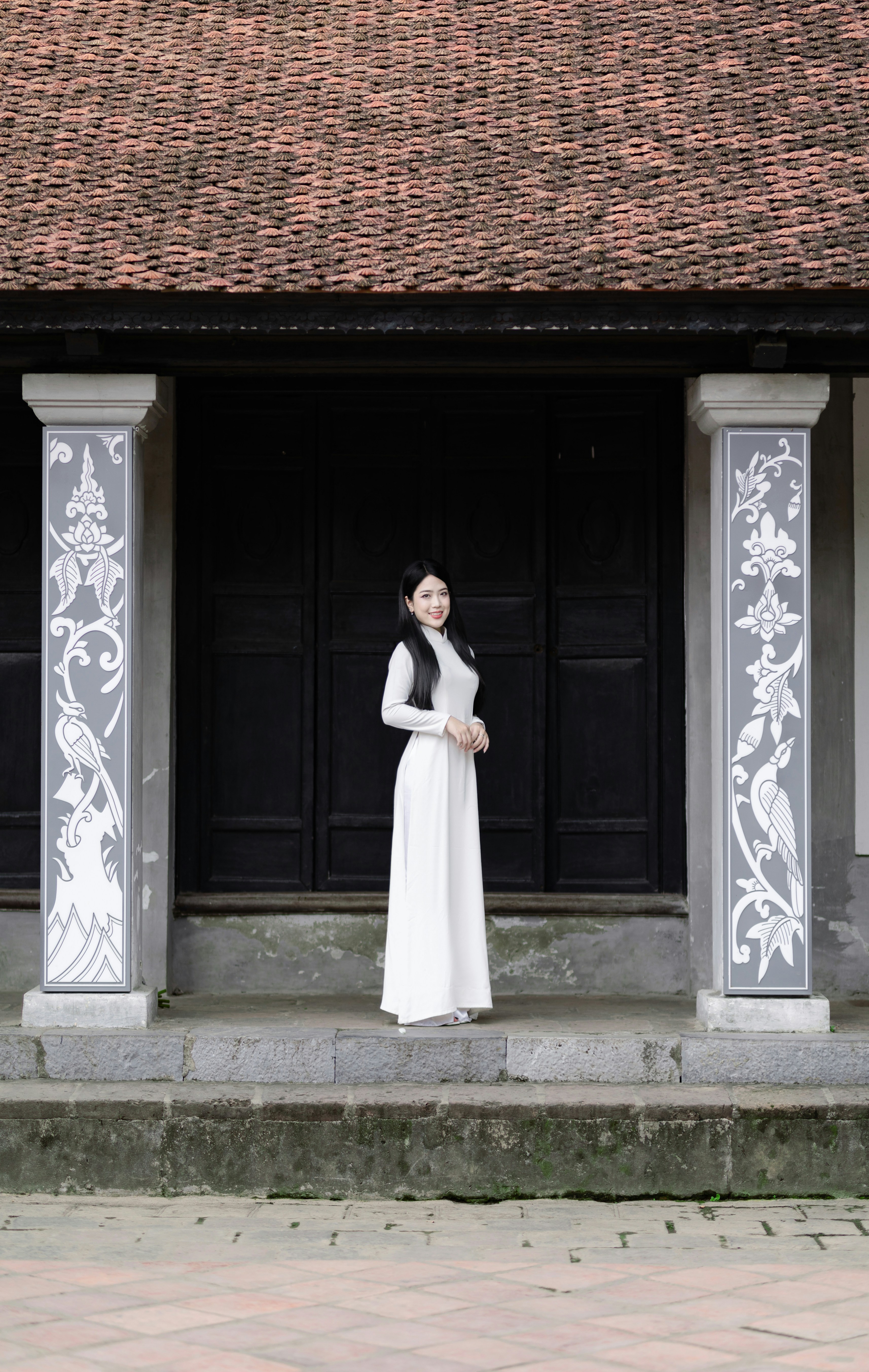 A woman in a traditional dress stands gracefully at the entrance of a historic building, accentuated by ornate columns and a textured roof.