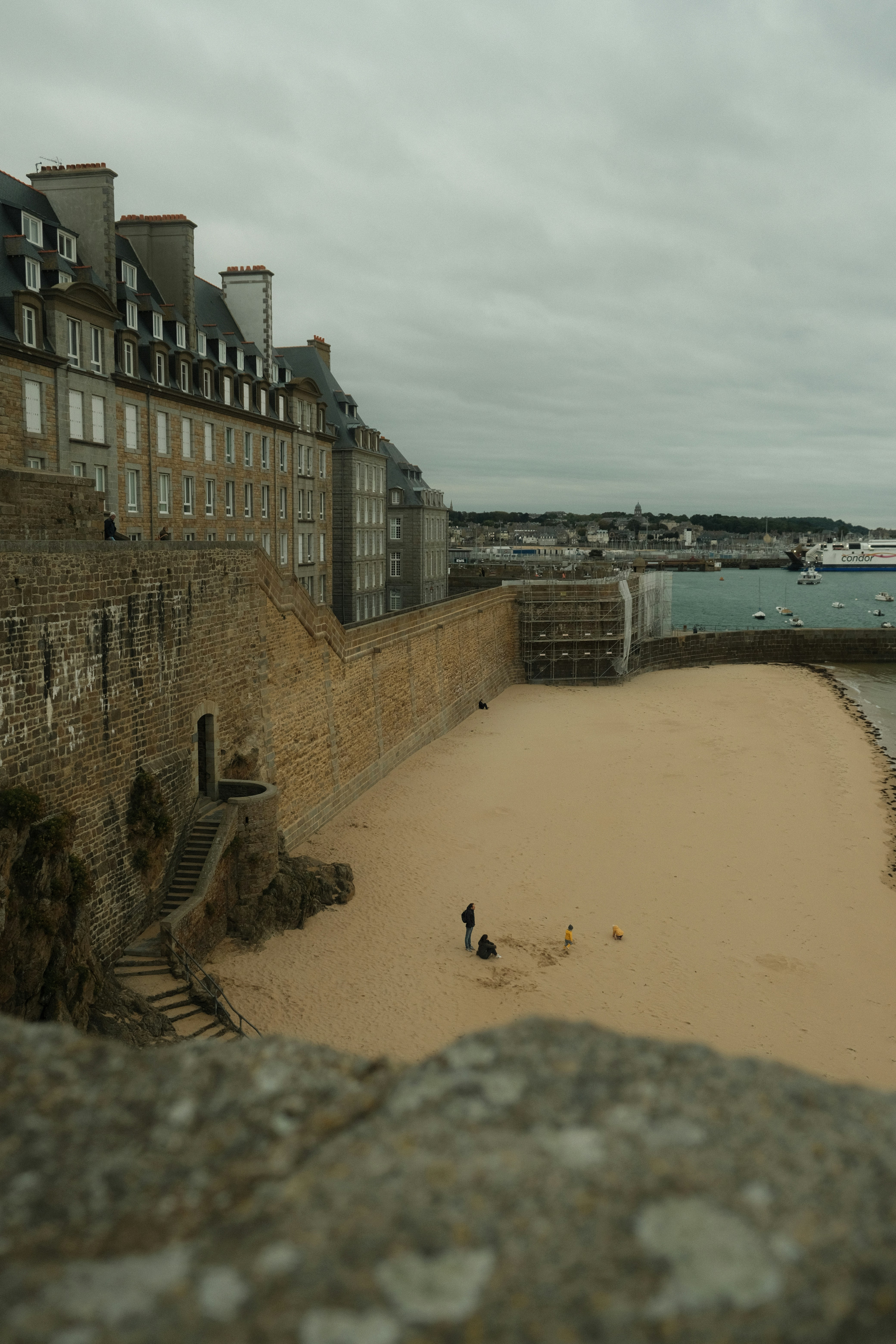 Historic stone walls embrace a serene beach, where two figures stroll along the sand near a tranquil harbor. 