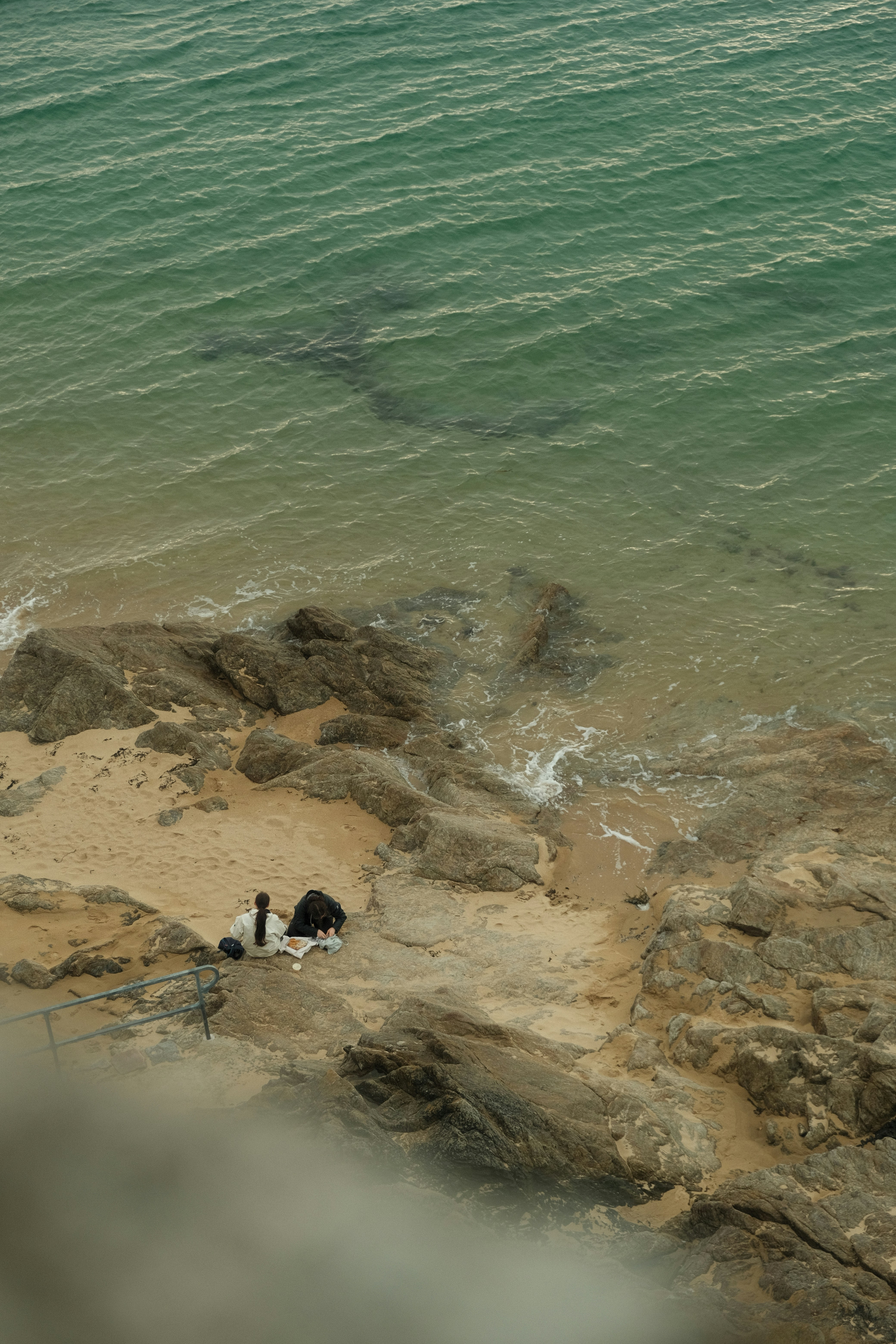 Two individuals seated on rocky beach, engaged in a thoughtful moment as gentle waves lap at the shore.