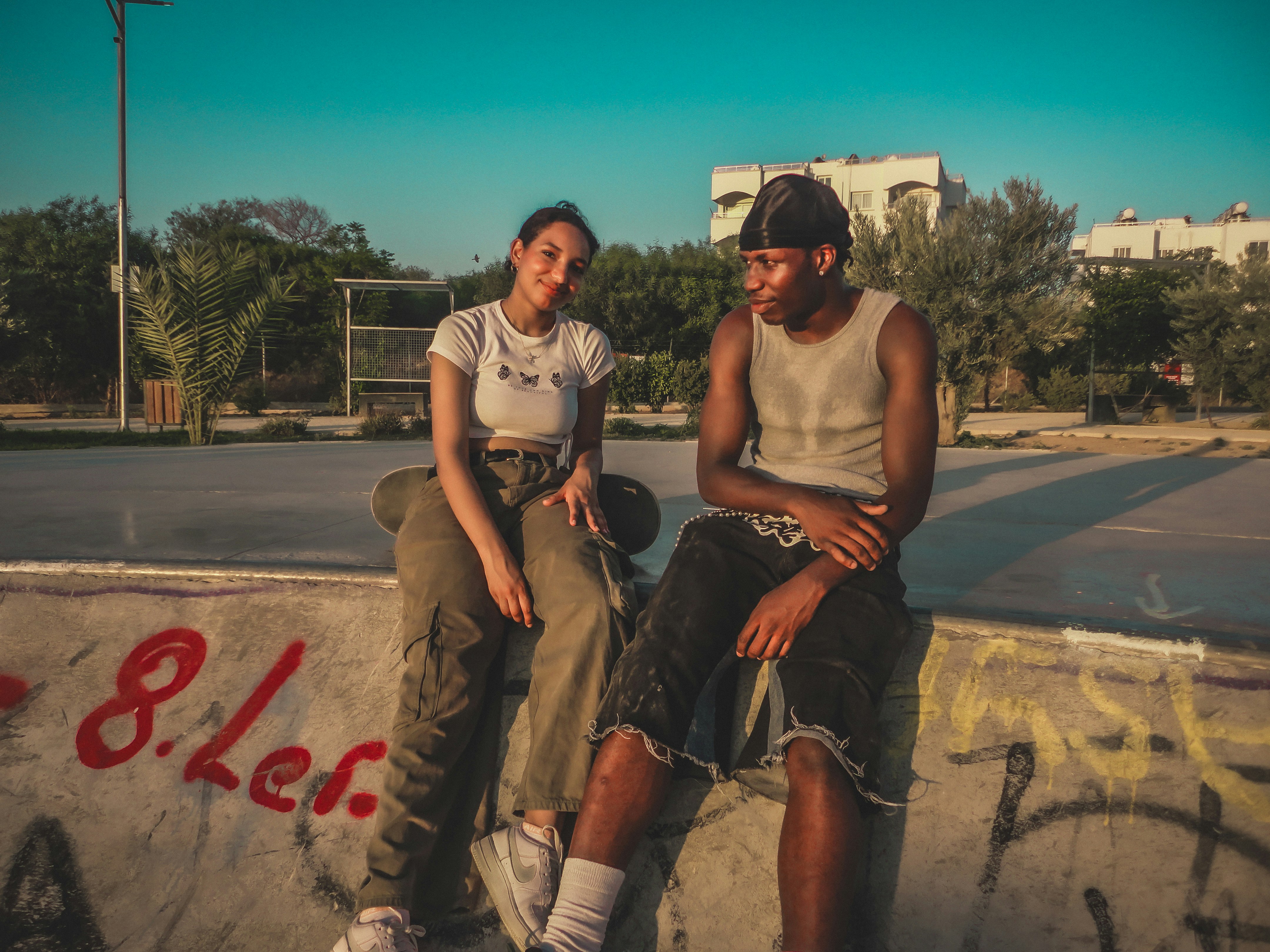 Two friends sitting on a skatepark ledge, enjoying a sunny day. The scene captures a moment of camaraderie and leisure.