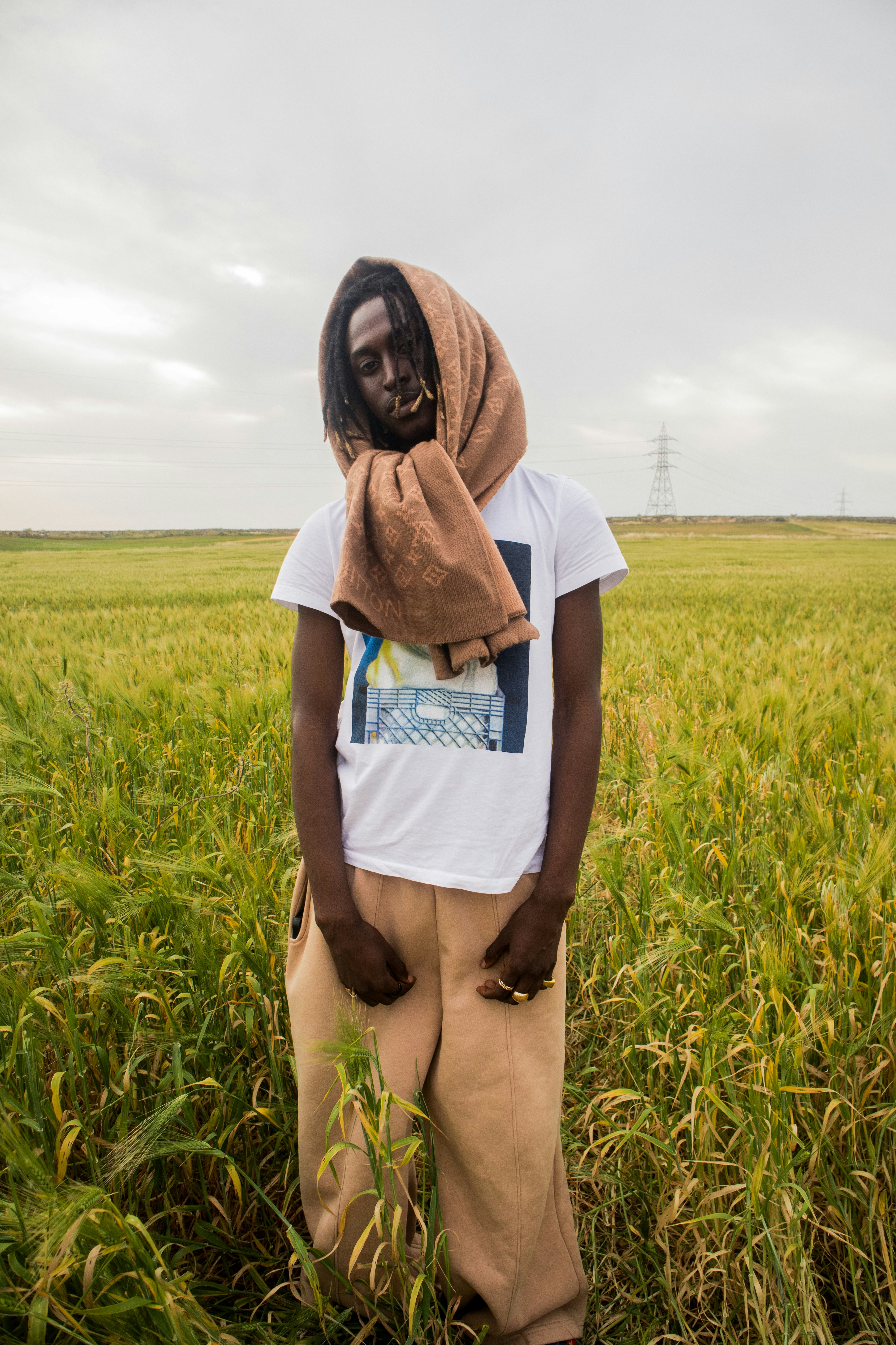 Individual standing in a field of green, adorned with a light brown scarf, exuding a serene connection with nature. The cloudy sky adds a moody backdrop.