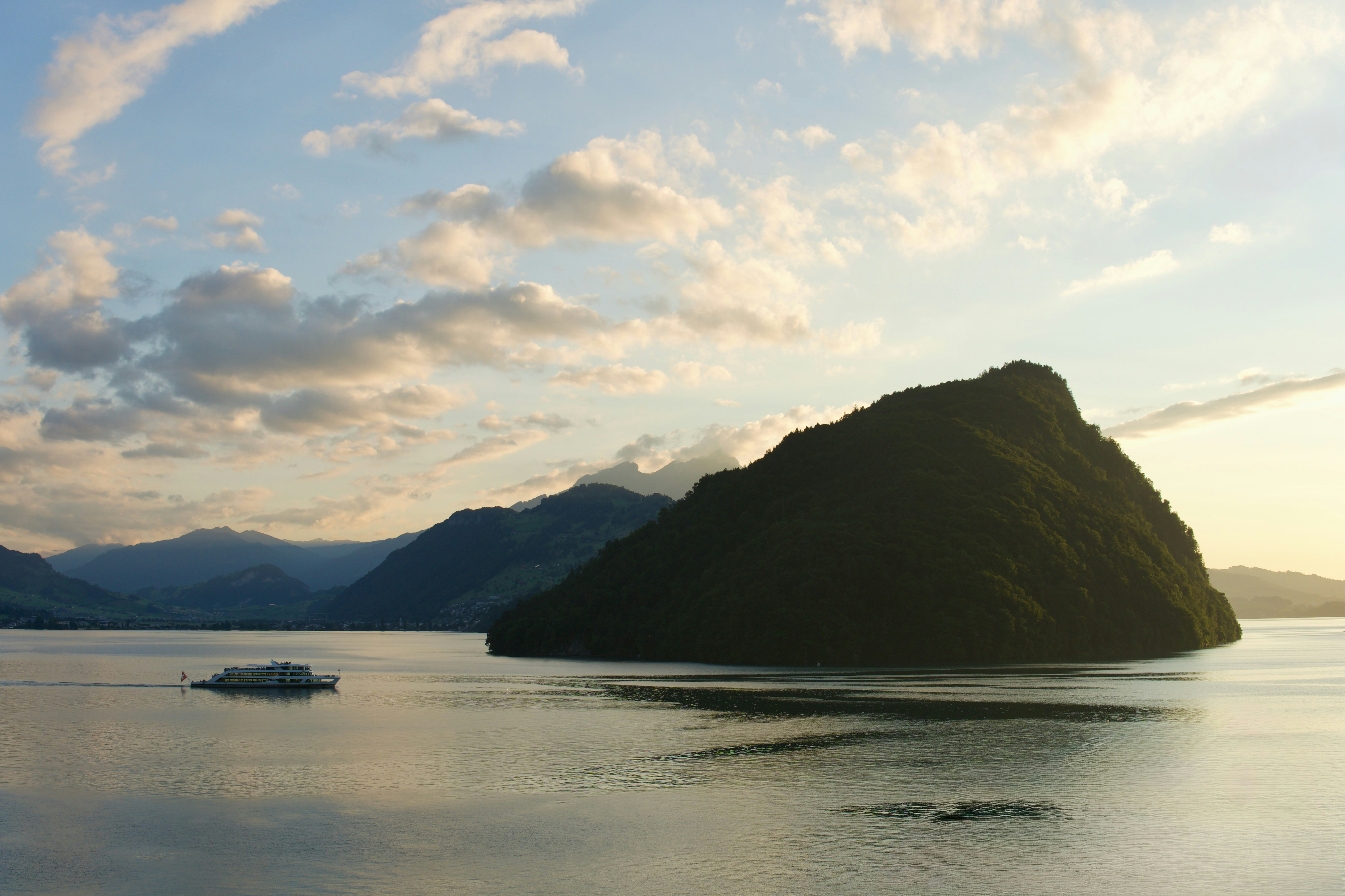 A tranquil lake scene featuring a lone boat gliding past a verdant island under a soft evening sky. The gentle ripples reflect the surrounding mountains.