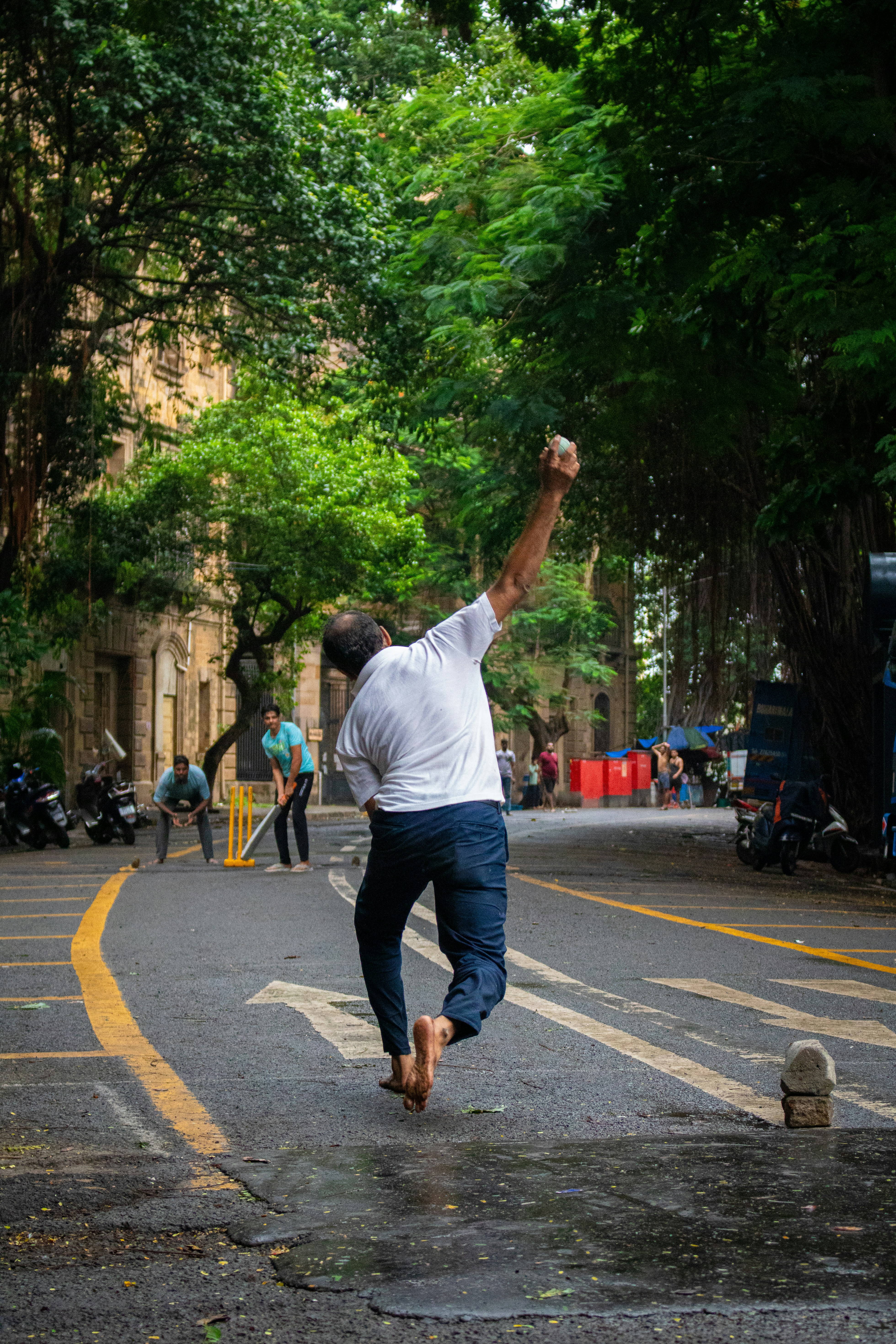 A group of men engaged in a lively game of street cricket on a quiet urban road, surrounded by lush greenery and historical architecture.