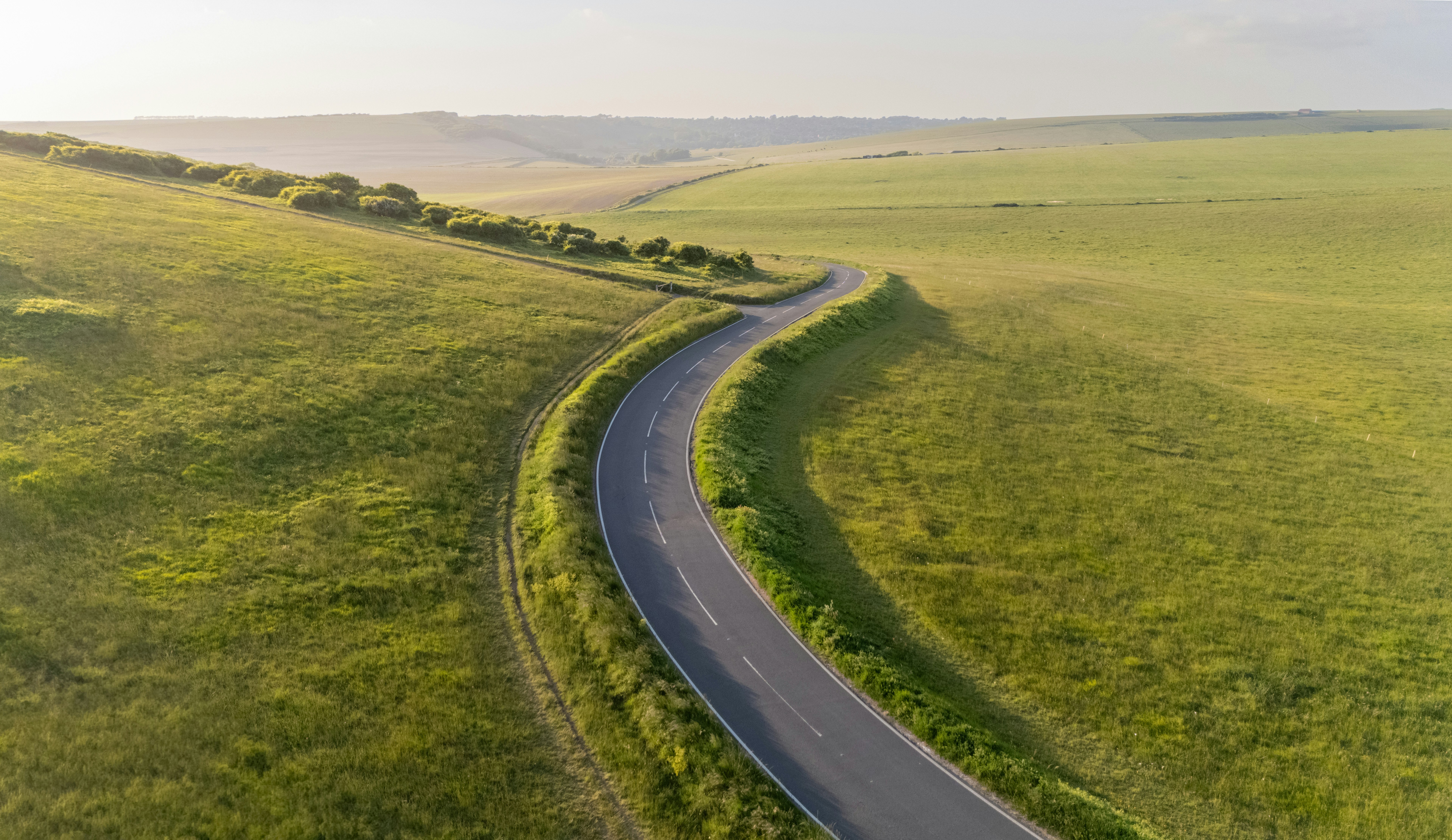 Curved road meandering through lush green fields under a soft sky. The landscape exudes tranquility and openness.