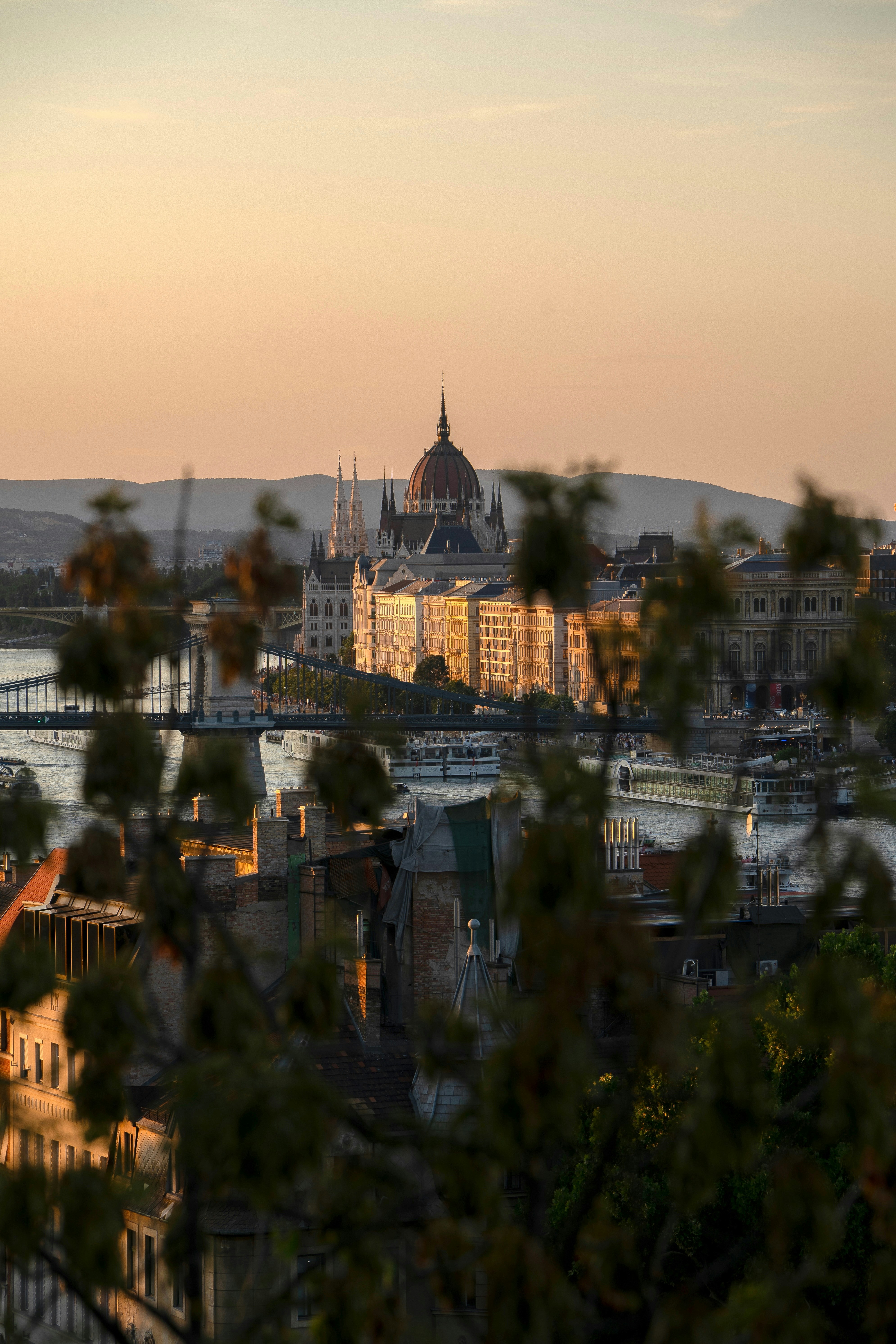 Budapest's iconic Parliament building bathed in golden sunset light, framed by foliage in the foreground. The serene Danube River reflects the warm hues of dusk.