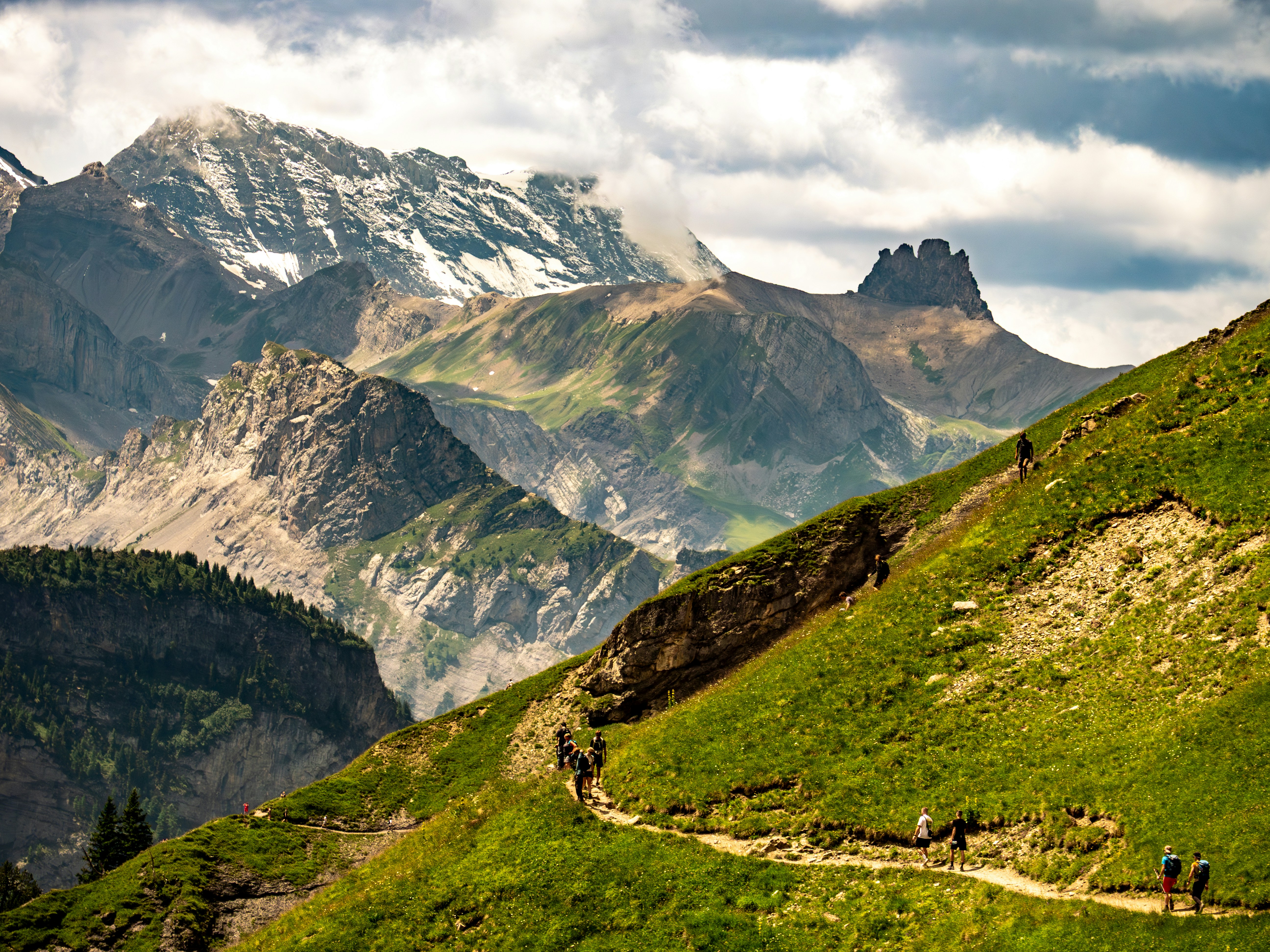 Hikers traverse a winding path through lush green hills, set against a backdrop of majestic mountains and dramatic skies.