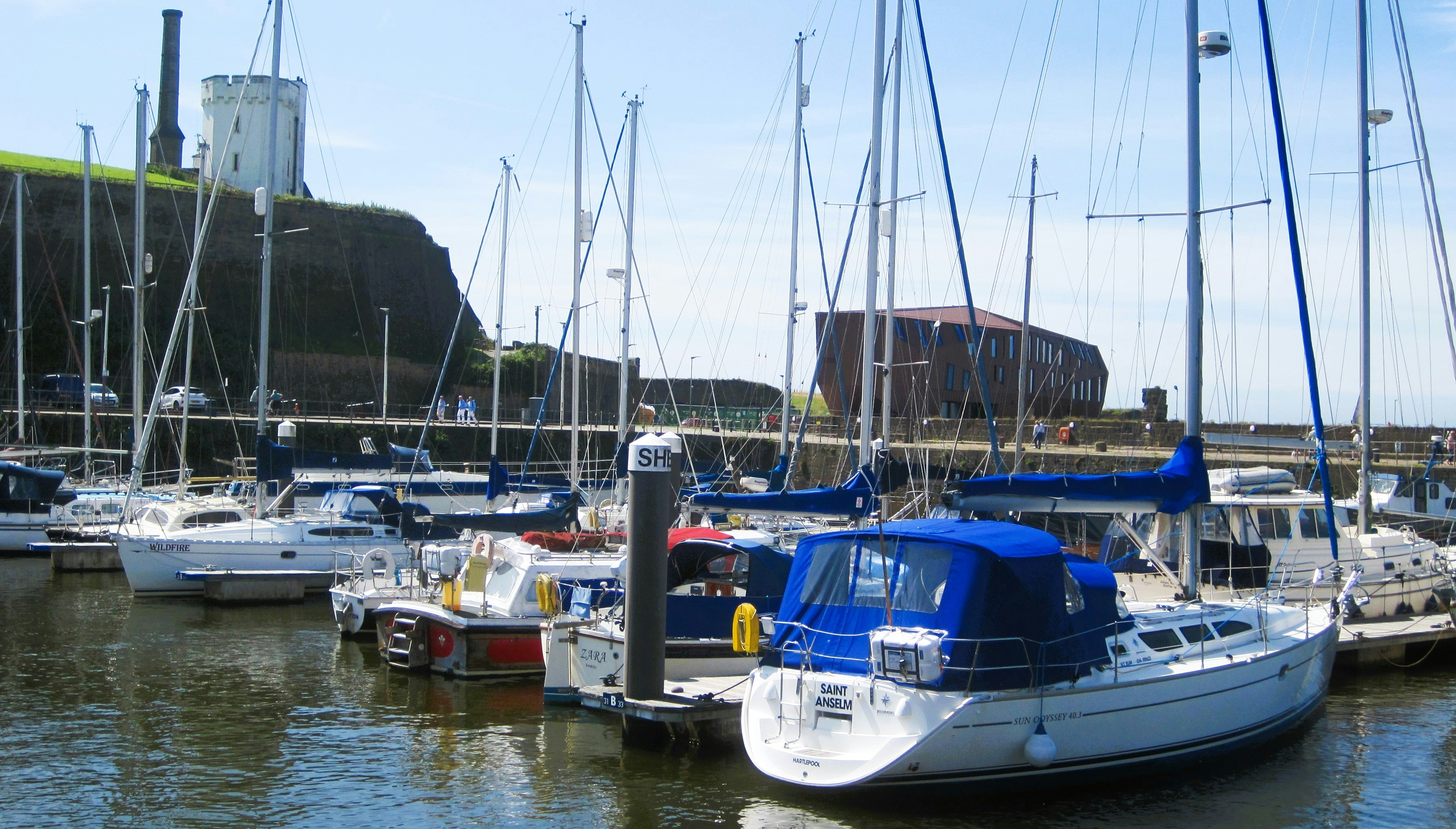 Sailboats docked in a serene marina, with a historic structure in the background and clear blue skies above.