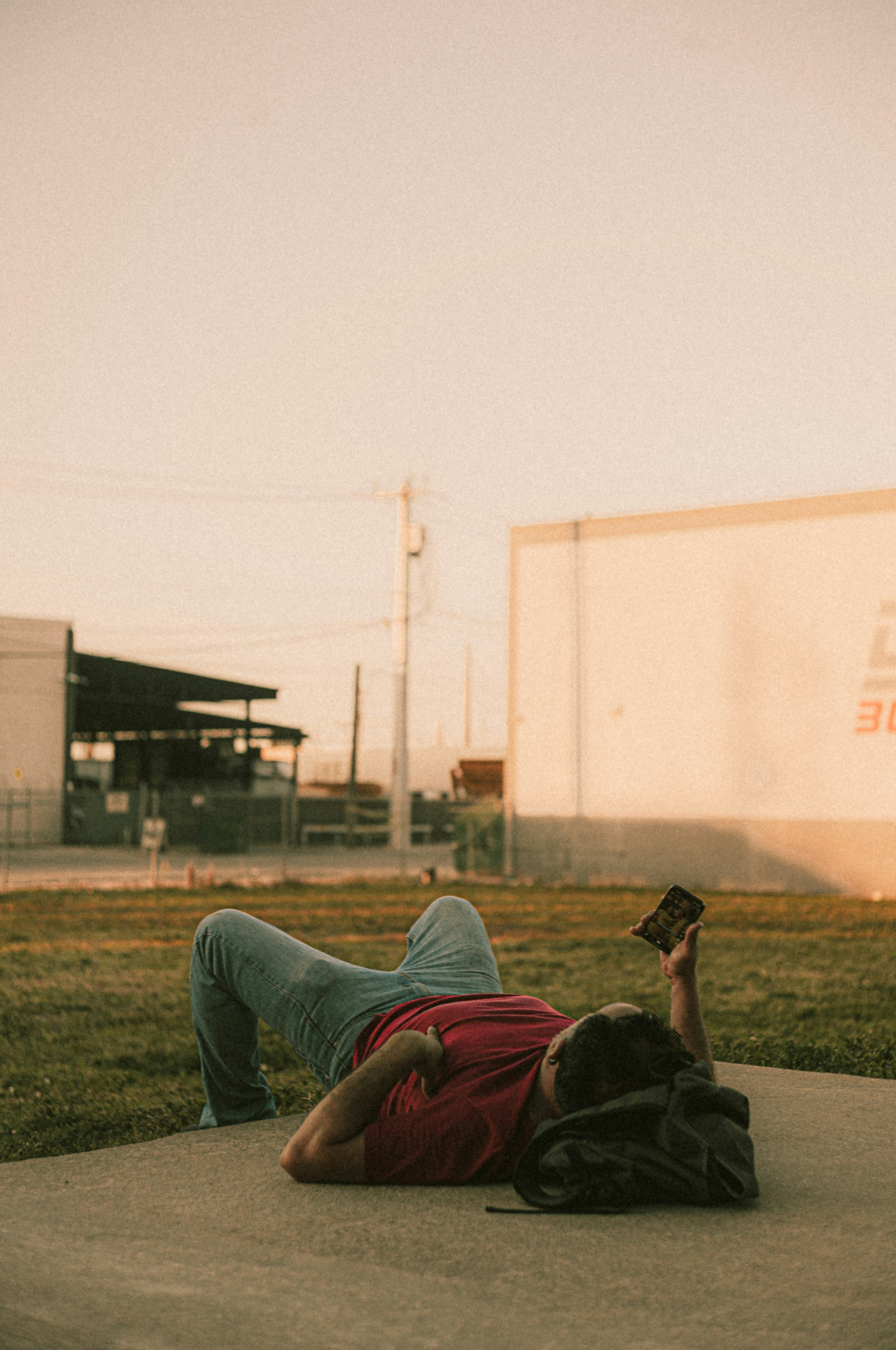 Individual reclining on a sidewalk, engrossed in a smartphone, surrounded by urban architecture and soft evening light.