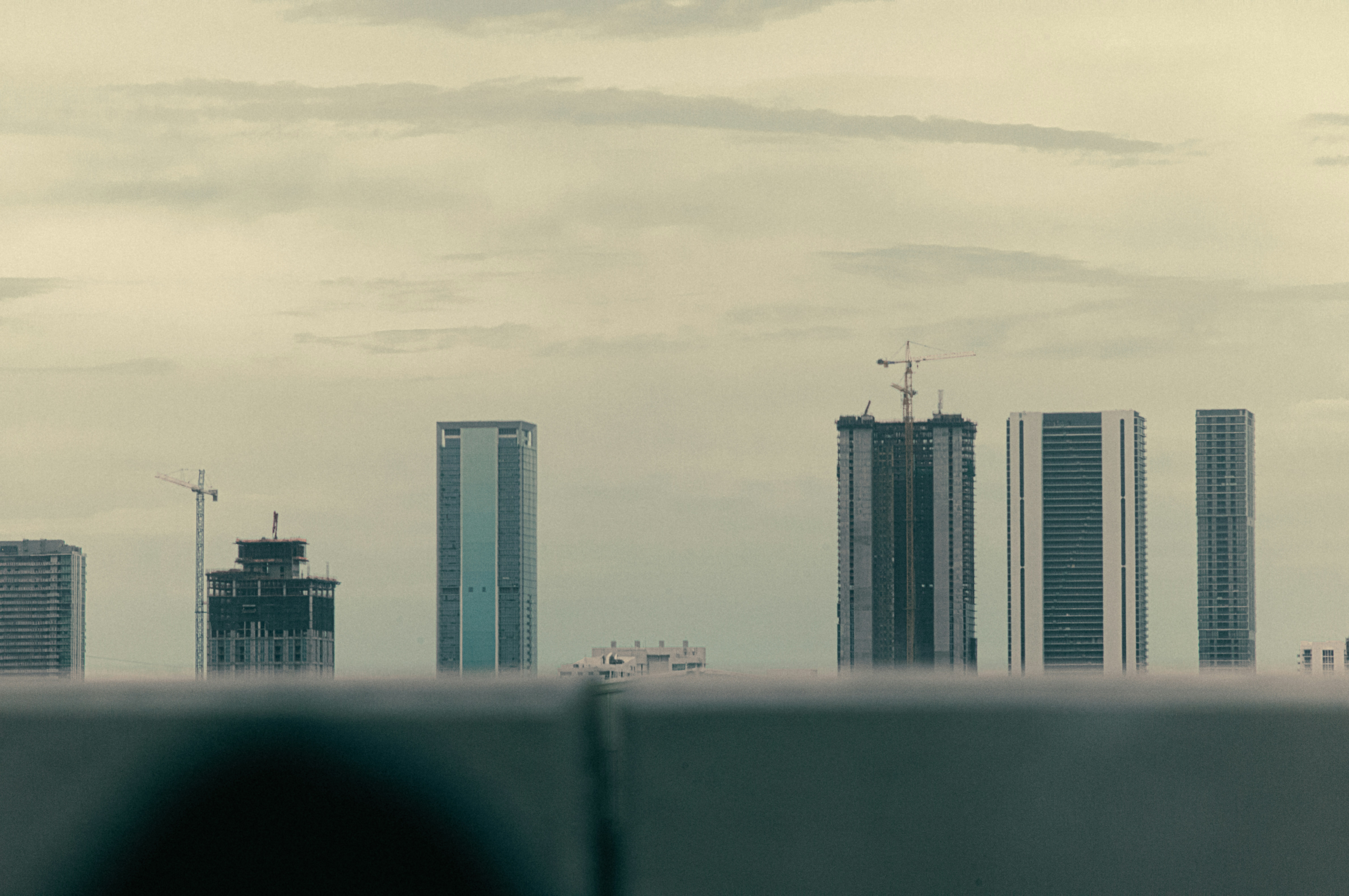 A skyline featuring a mix of modern skyscrapers and construction cranes under an overcast sky, emphasizing urban development.