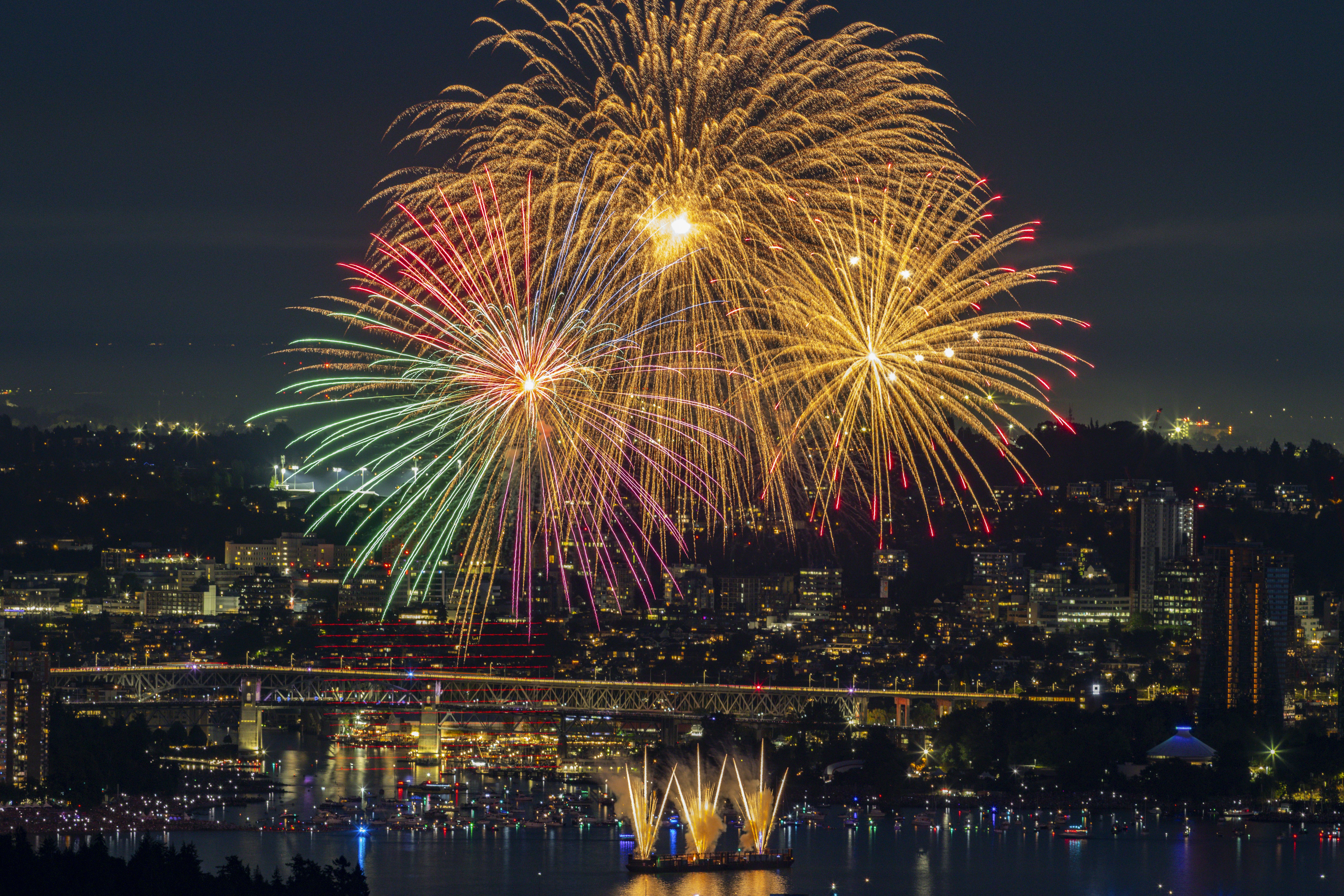 Vibrant fireworks burst across the night sky, illuminating a city skyline reflected in the water below.