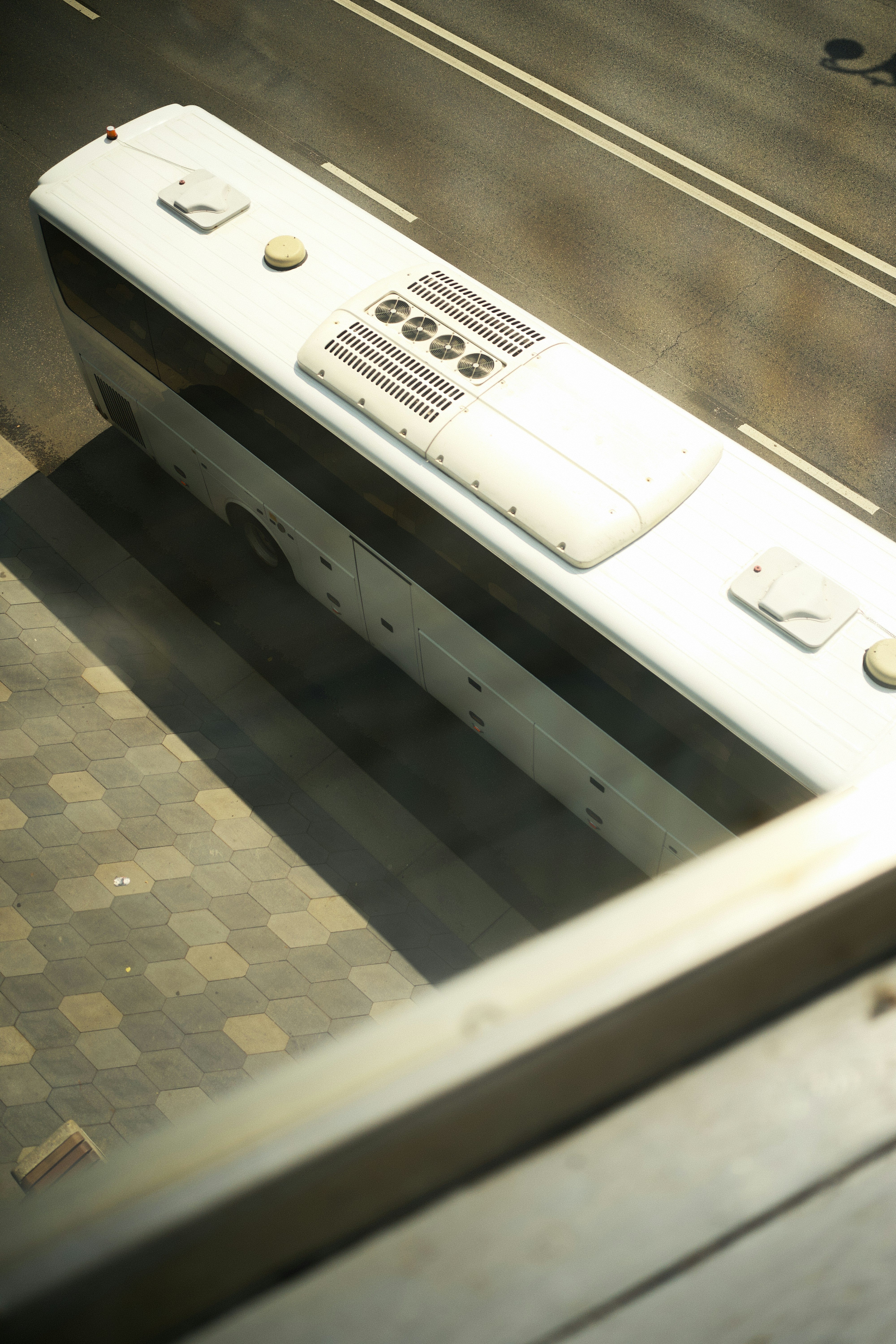 A white bus viewed from an elevated perspective, parked on a patterned sidewalk under natural light. The composition highlights the contrast between the bus and its surroundings.