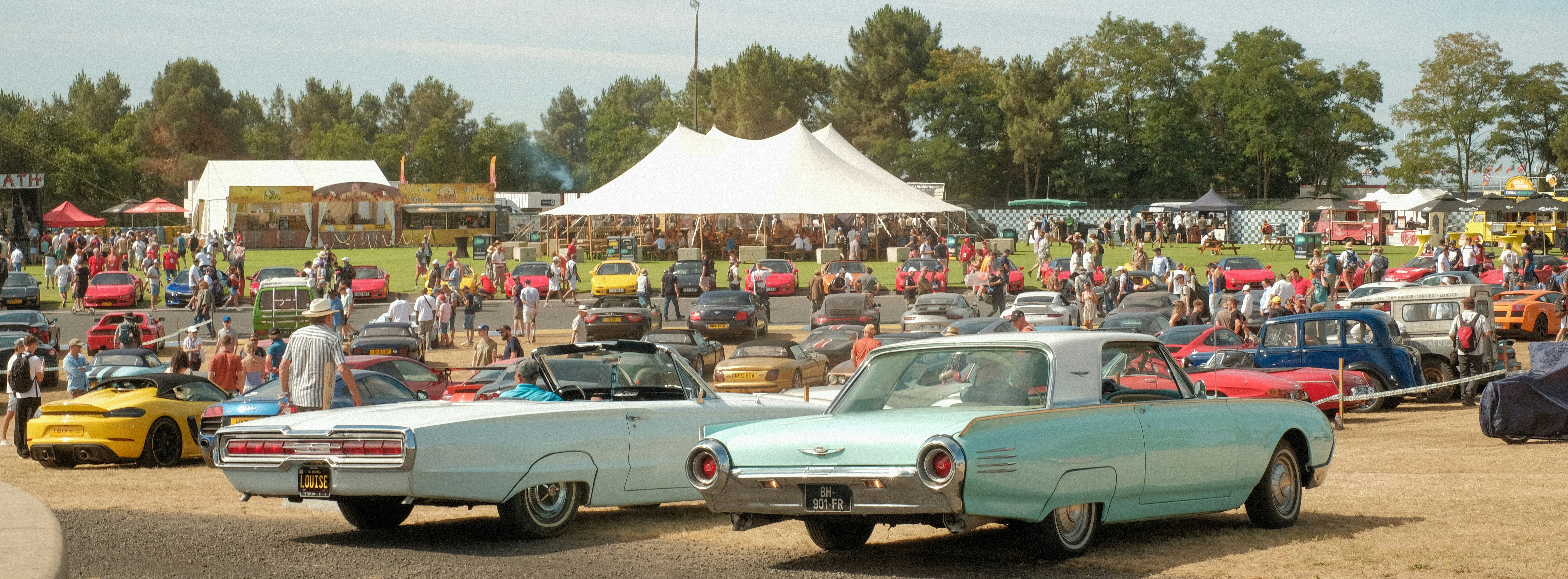 Two vintage cars parked in the foreground with a bustling car show in the background, showcasing an array of classic vehicles and enthusiastic attendees.