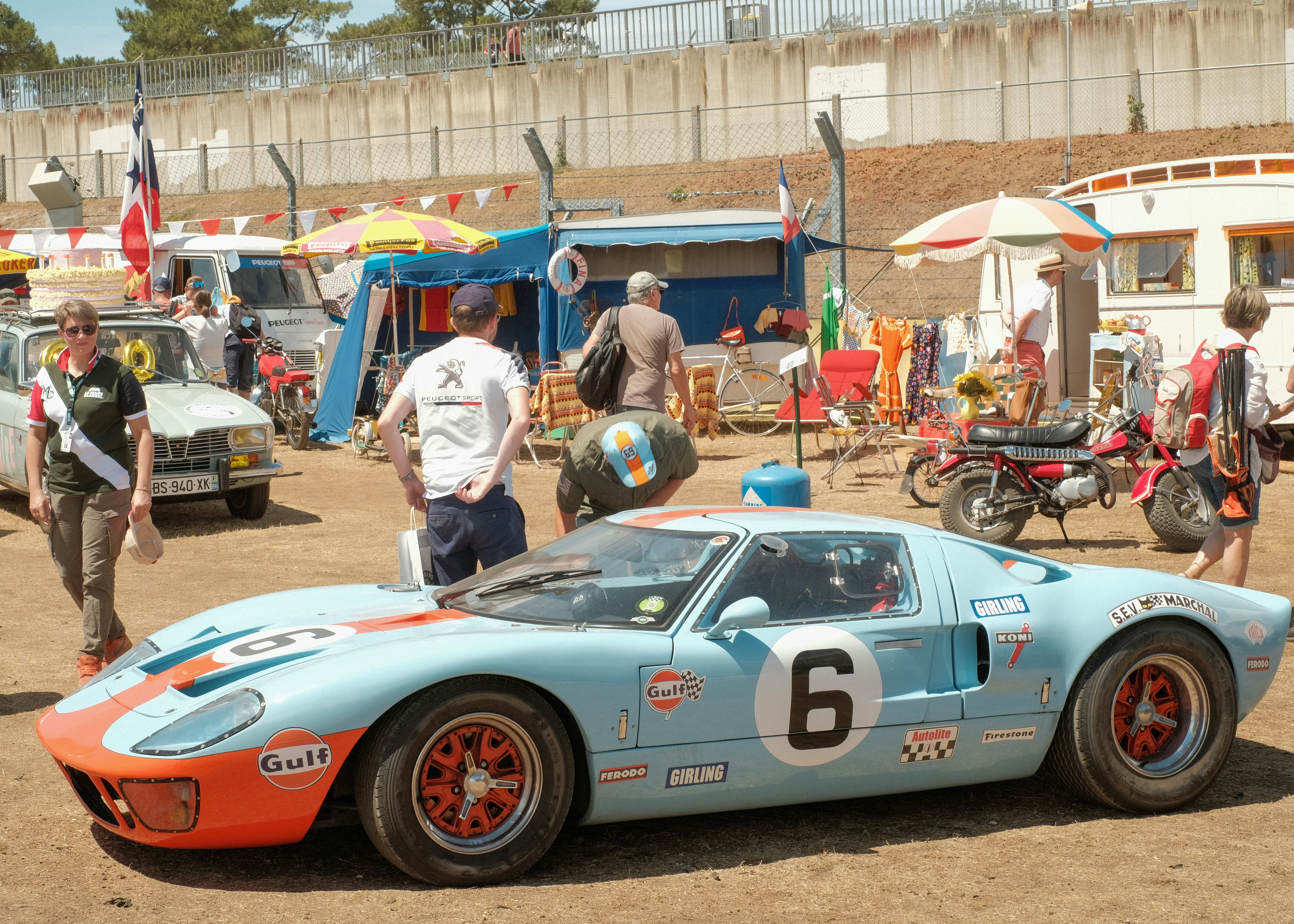 A classic Gulf-liveried Ford GT40 parked amidst a lively vintage racing event, surrounded by spectators and colorful tents.