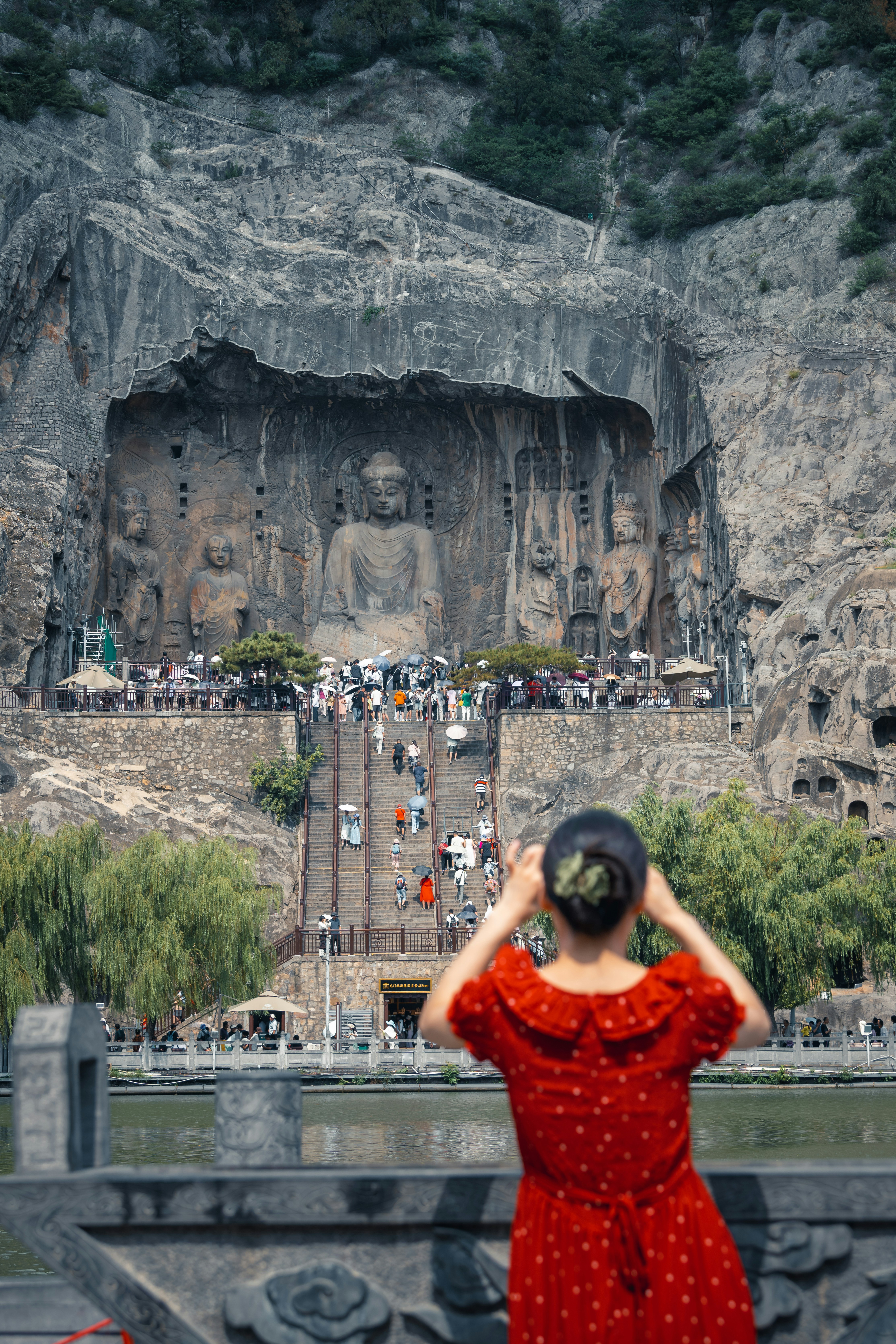 A woman in a red dress gazes at the Longmen Grottoes, featuring intricate Buddhist sculptures carved into the cliffside, surrounded by visitors and lush greenery.
