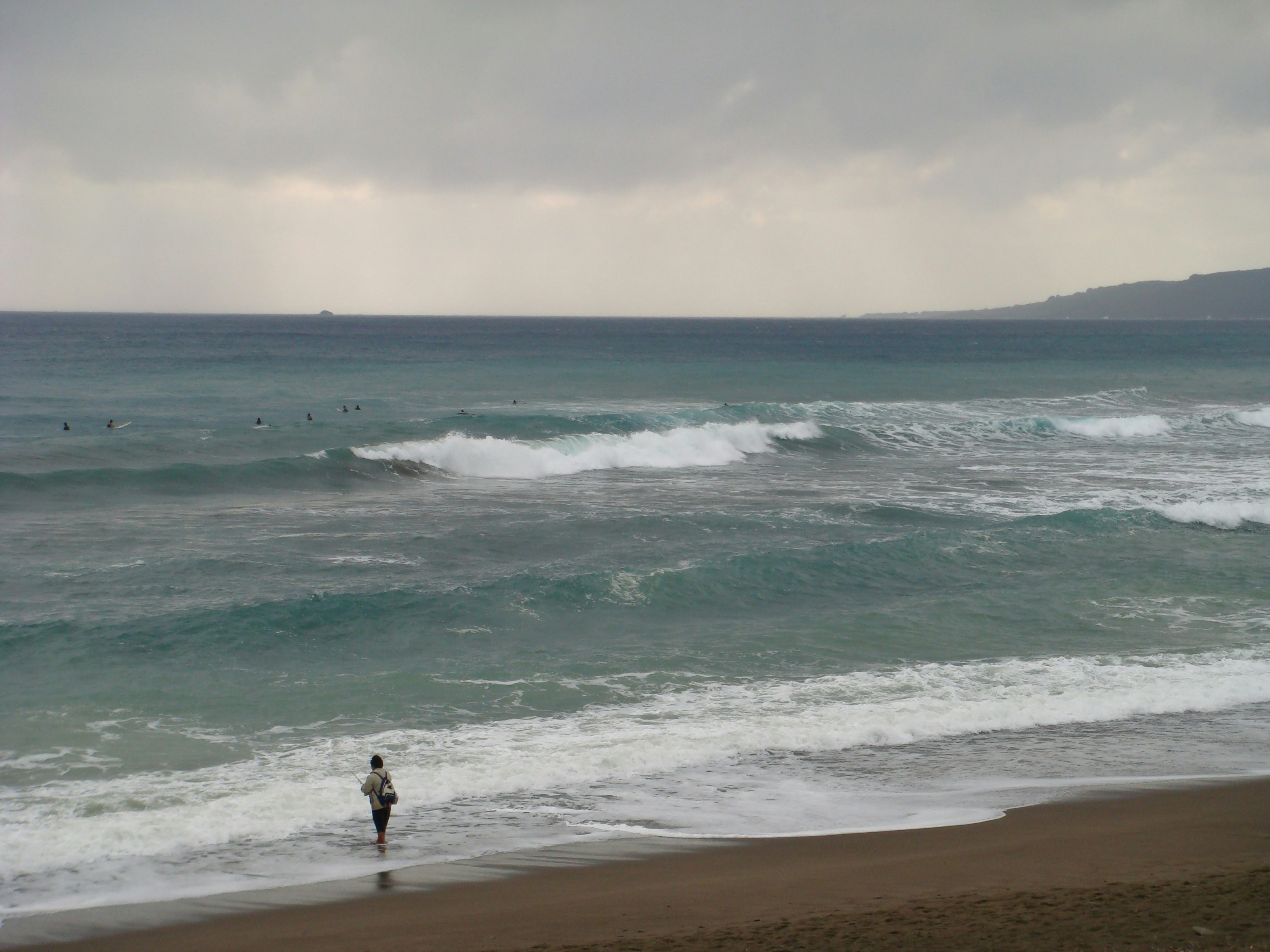 A lone figure wades along the shoreline, waves gently crashing against the sand under a moody sky. Surfers dot the distant waters, embracing the surf.