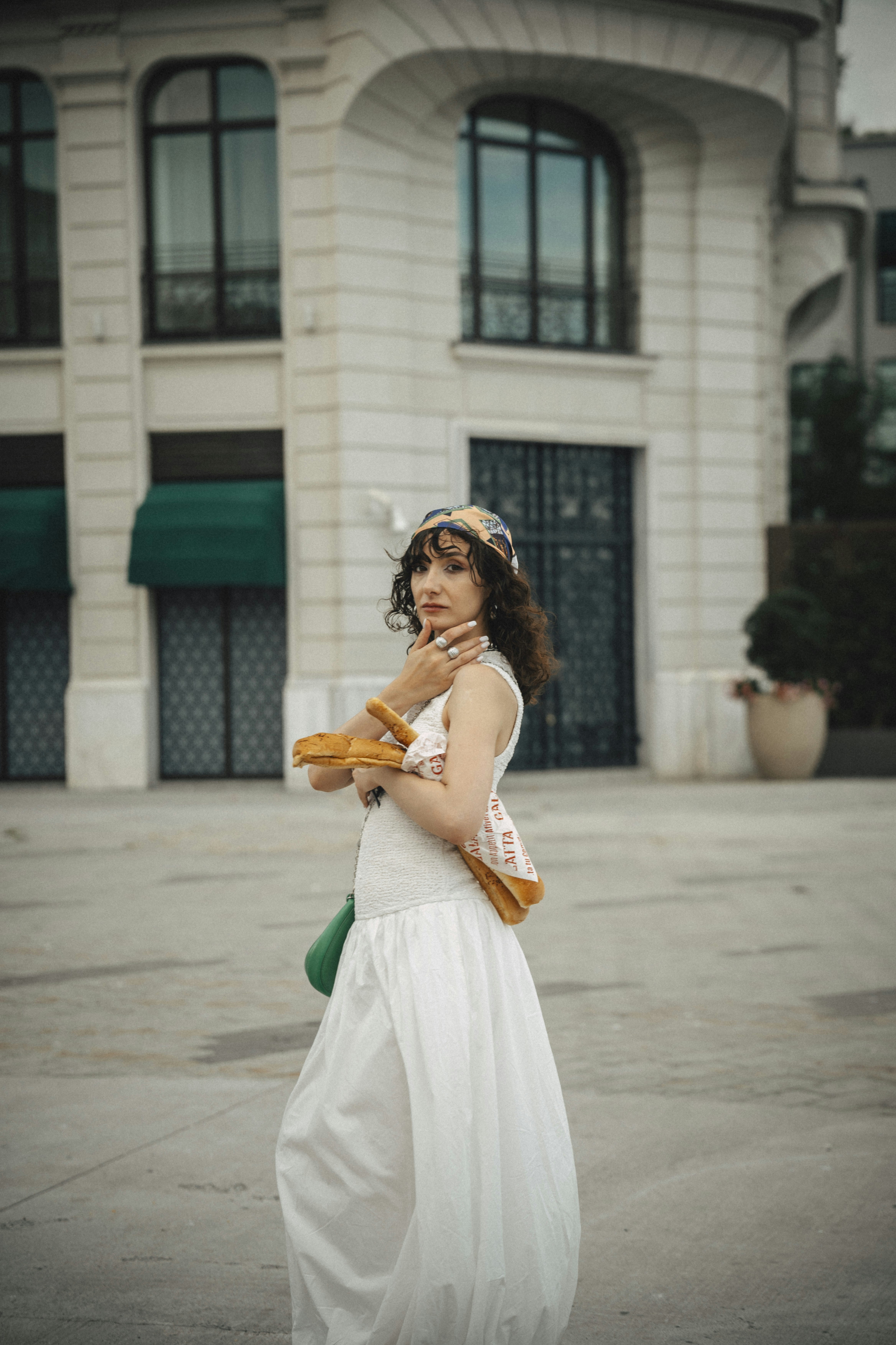 A woman in a flowing white dress poses confidently, holding a vibrant yellow accessory against an elegant urban backdrop.