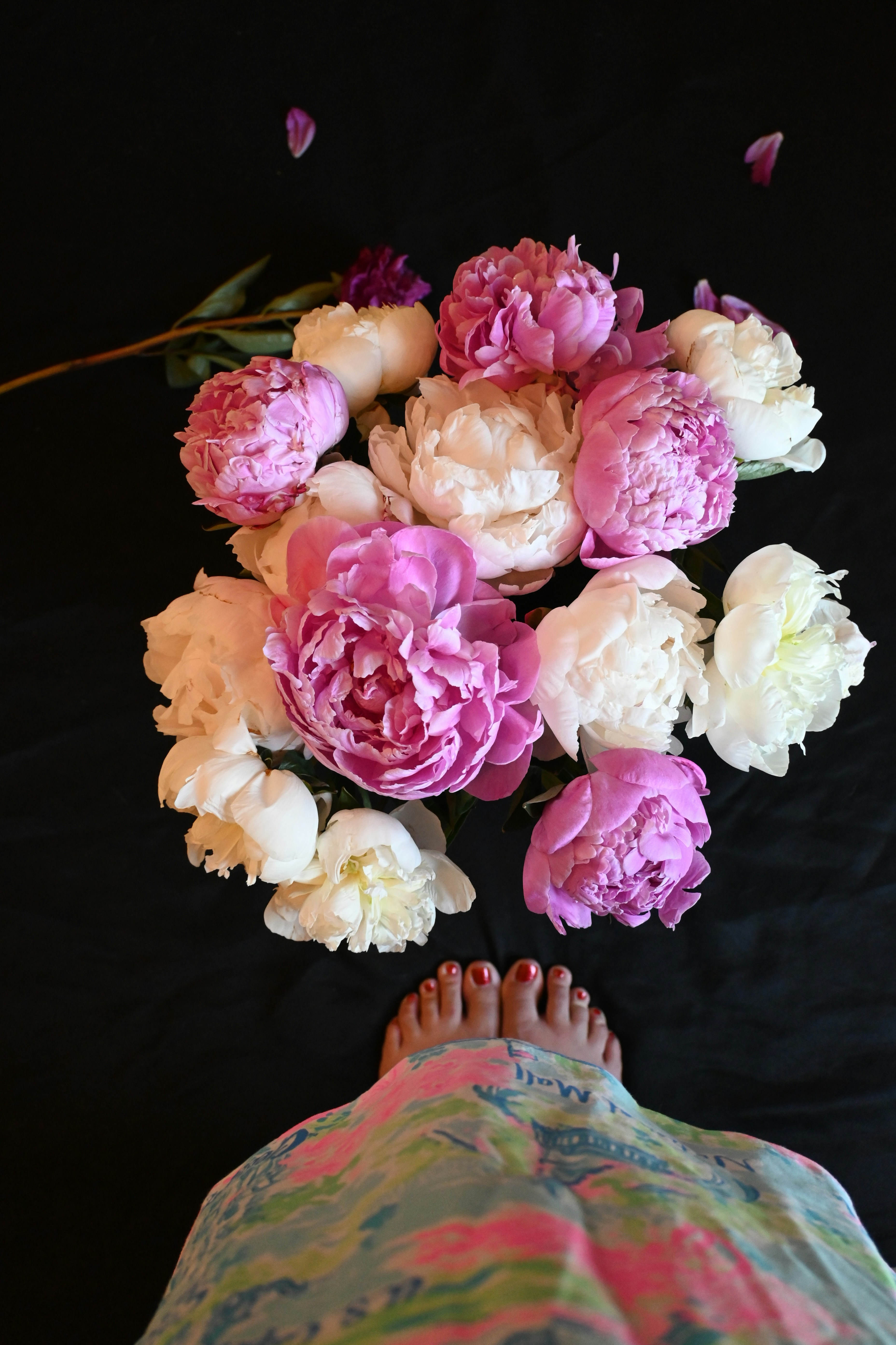 A vibrant arrangement of peonies in various shades atop a dark background, with a person's feet in a colorful dress positioned below. 