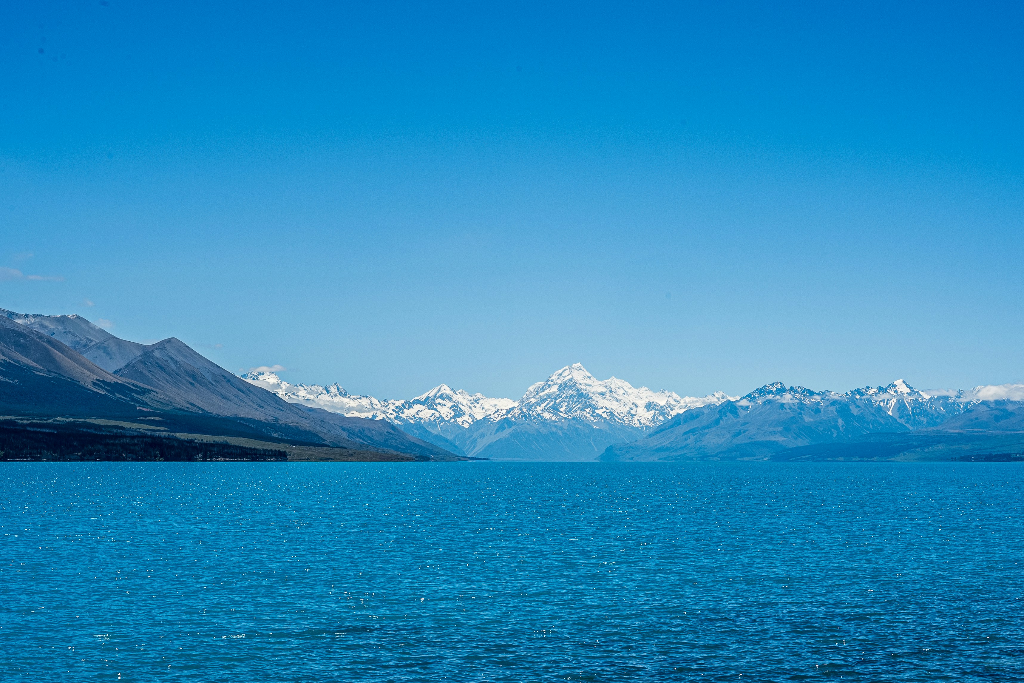 Snow-capped mountains reflect in the tranquil waters of a vast lake under a clear blue sky.