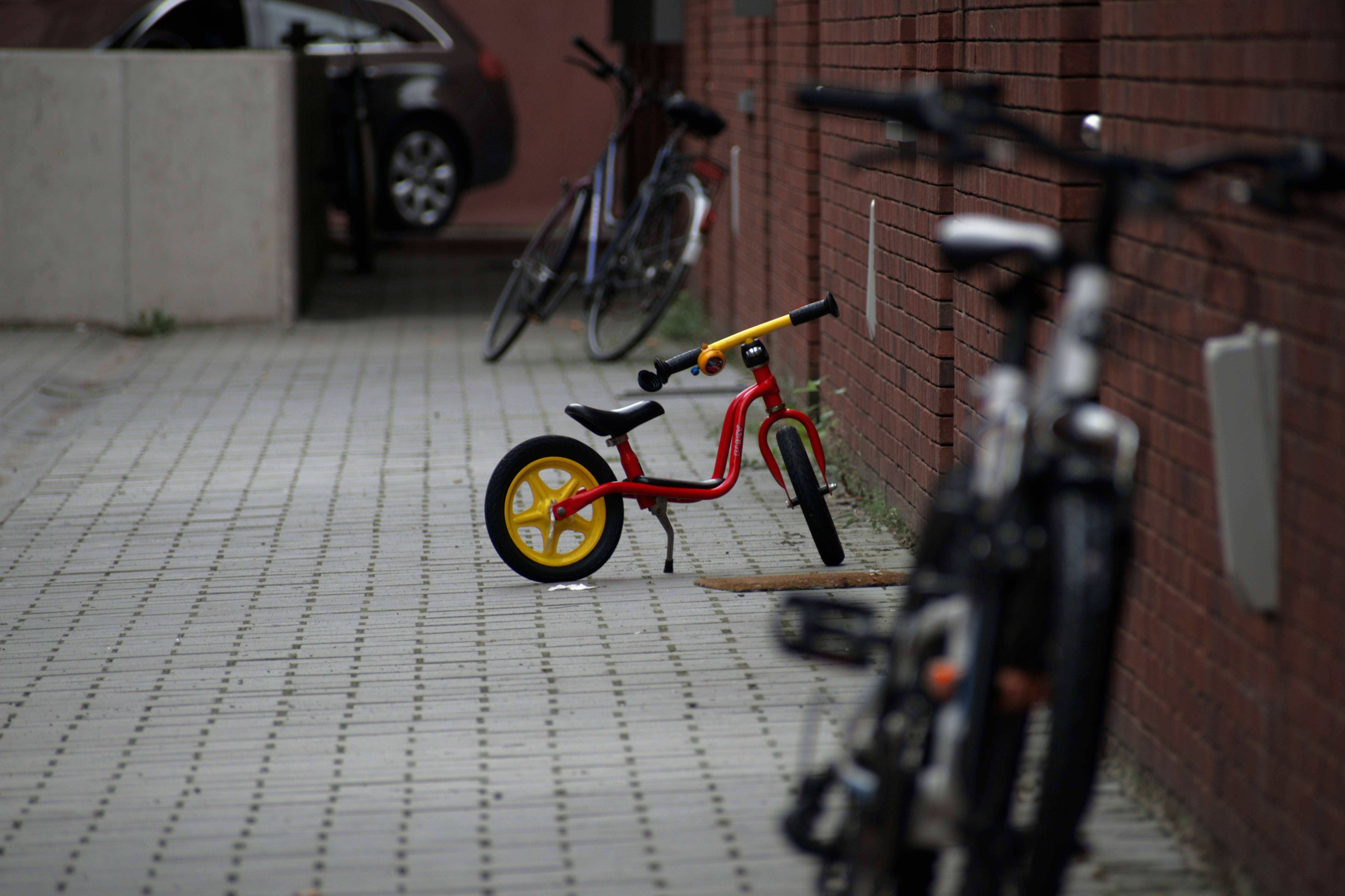 Bright red and yellow tricycle stands alone on a cobblestone path, surrounded by parked bicycles and a muted urban backdrop.