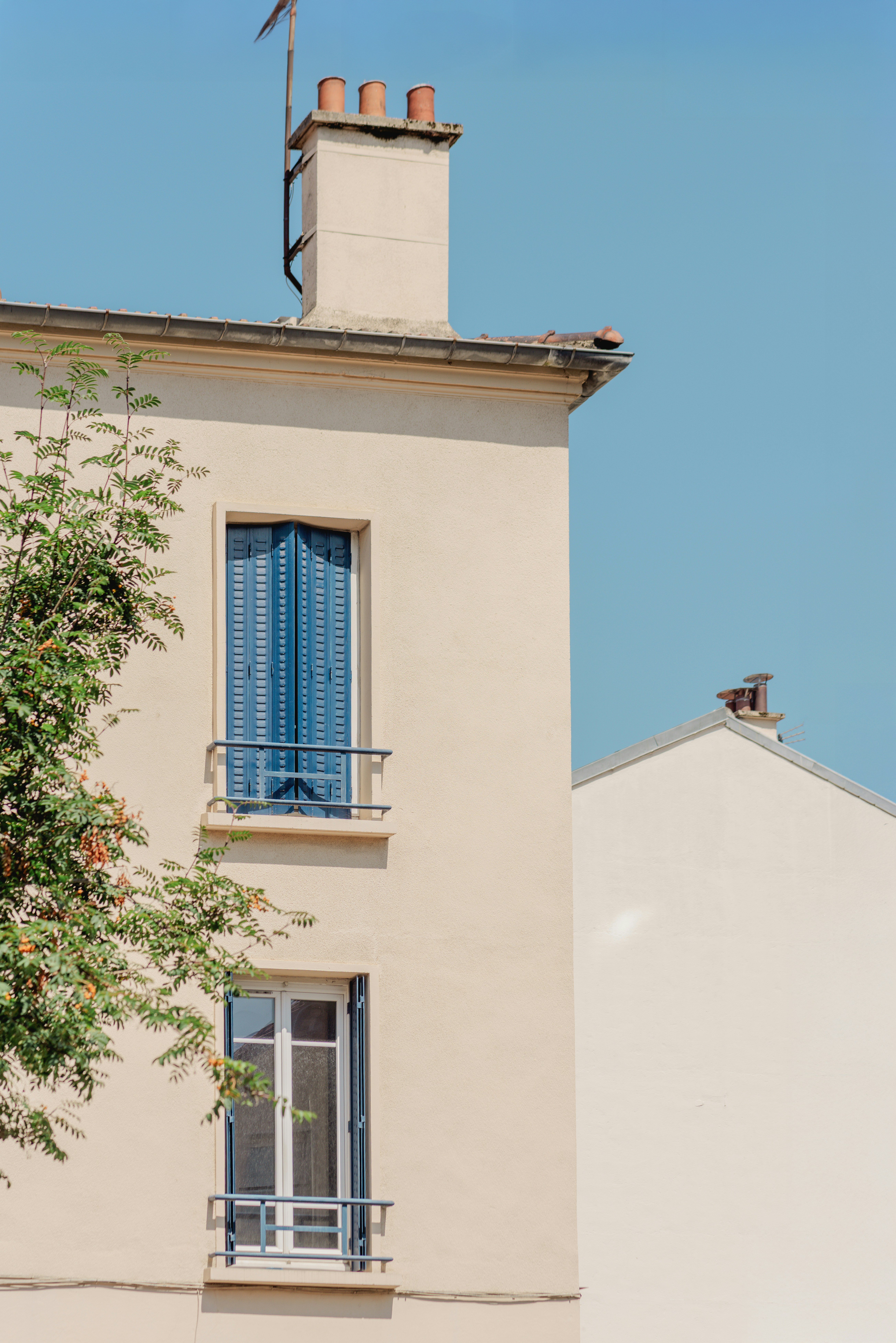 Light-colored building facade featuring a blue window with shutters, framed by lush greenery. A contrasting white wall completes the urban scene.