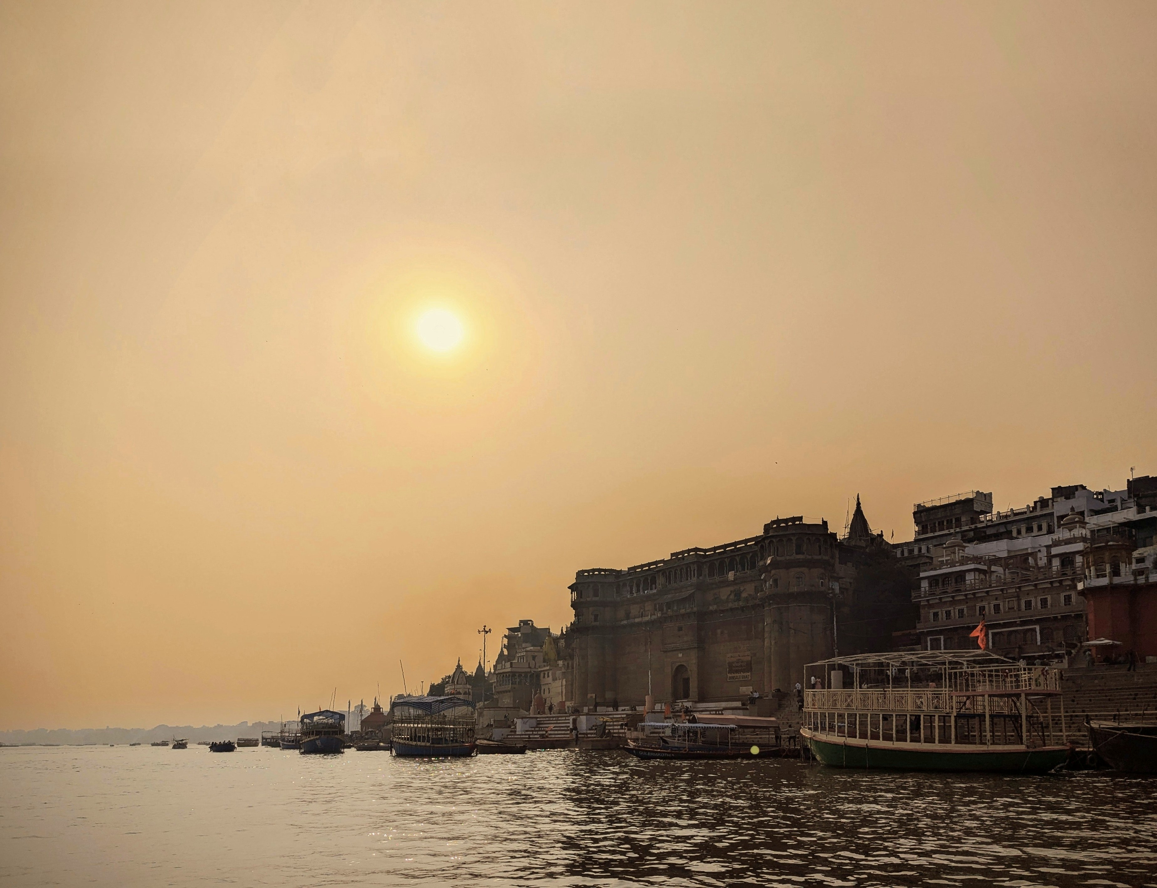 Silhouetted boats line the banks of the Ganges as the sun sets behind ancient architecture, casting a warm glow over the water.