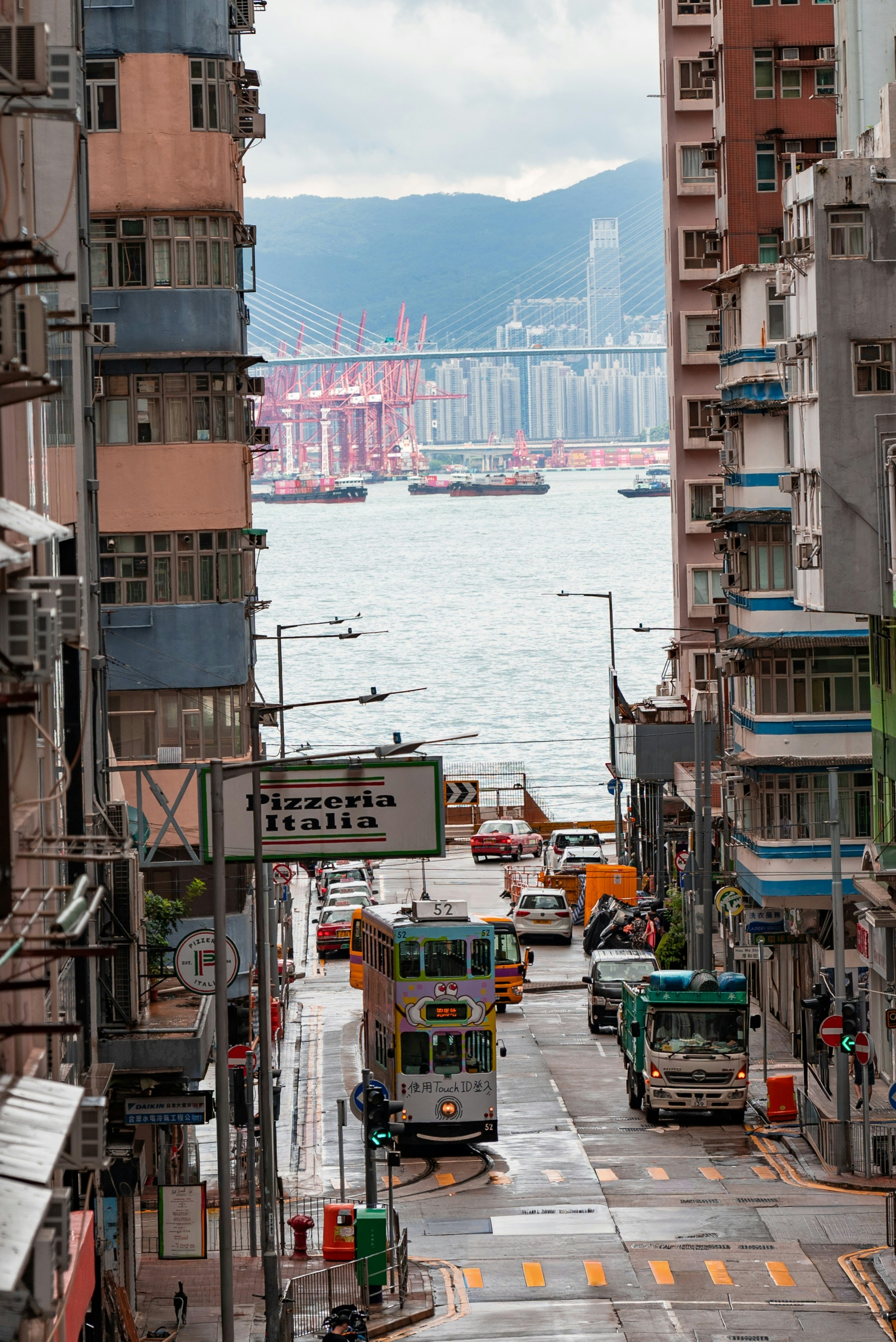 A bustling street scene in Hong Kong showcasing a tram navigating through the city, with harbor views and colorful buildings lining the road.