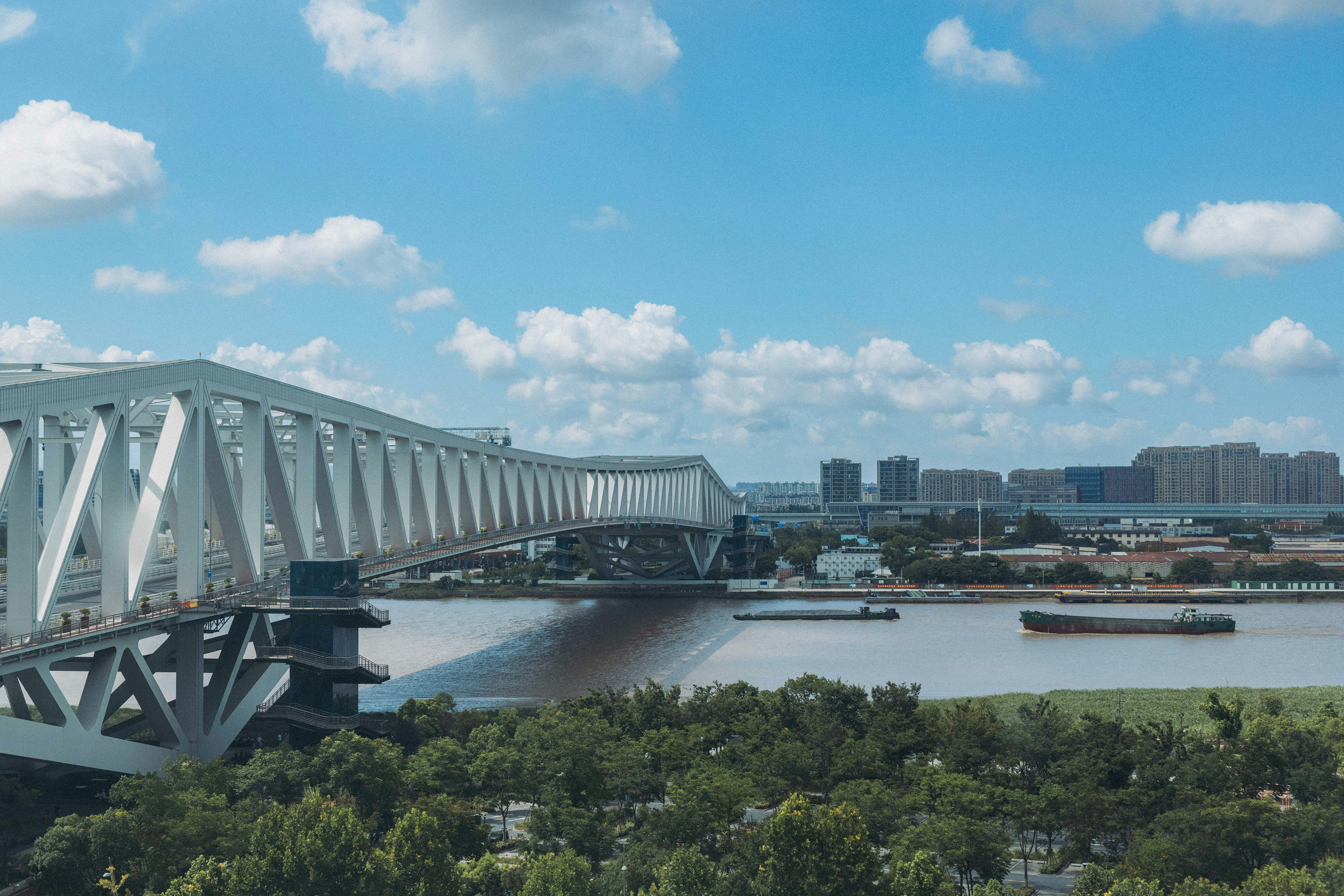 Modern white bridge spanning a river, with cargo ships navigating the water and urban structures in the background. Lush greenery frames the scene.