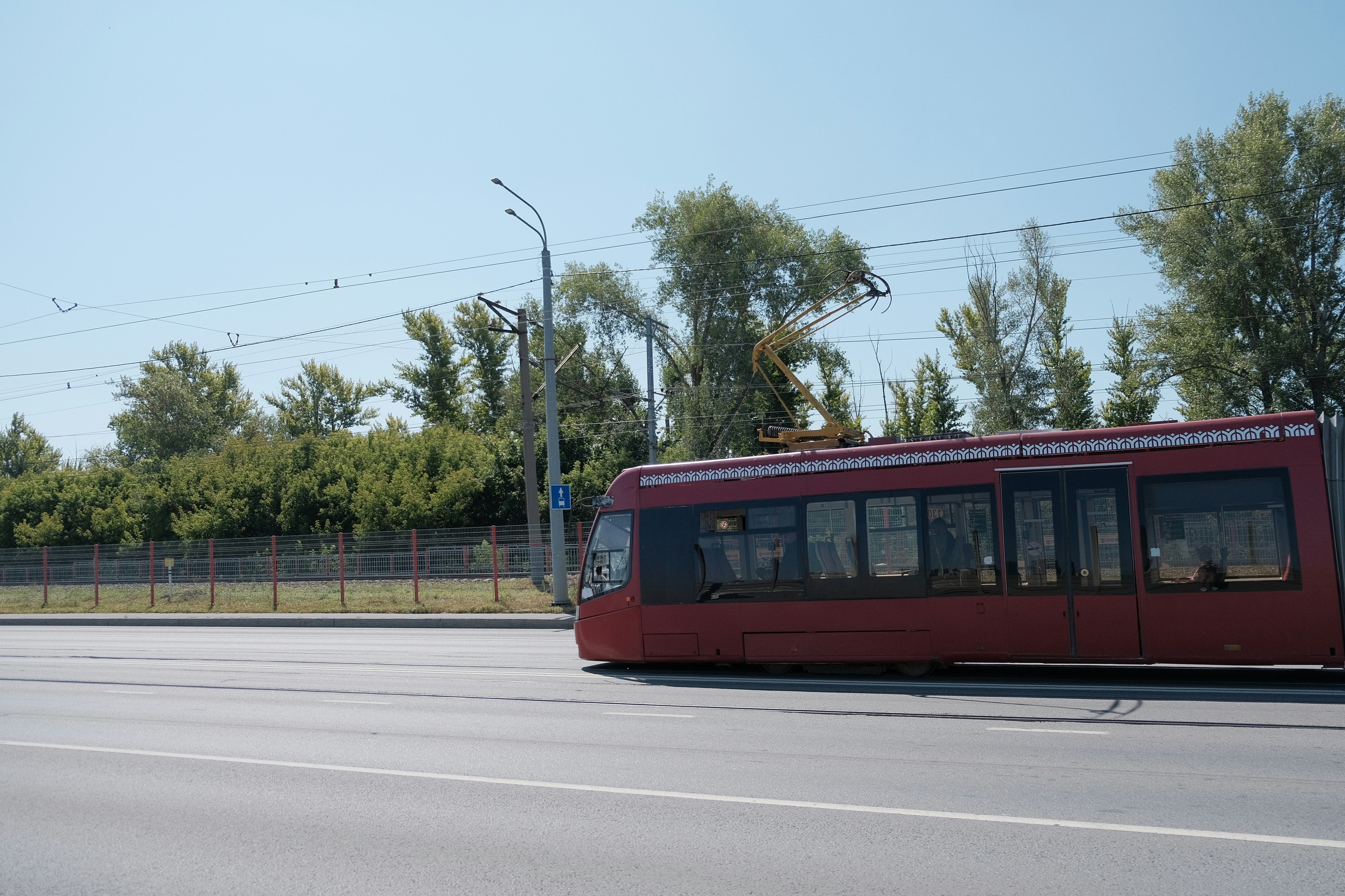 A red tram with decorative patterns is parked on a street, surrounded by trees and utility poles under a clear blue sky. | A red tram is moving along a road.
