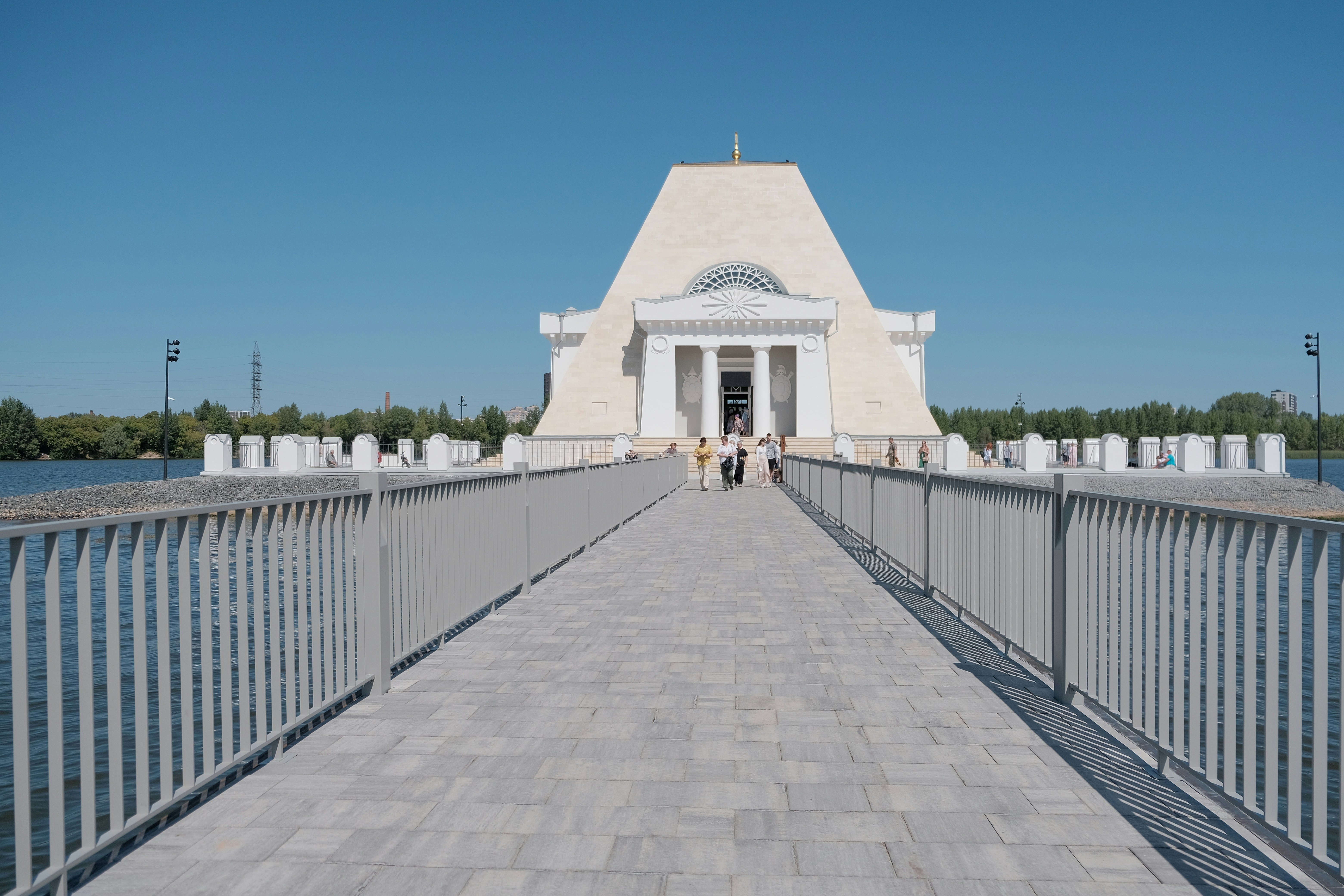 A white, pyramid-shaped monument with classical architectural details stands near a body of water, surrounded by people walking along a paved pathway under a clear blue sky. | A temple-like building stands on the water.