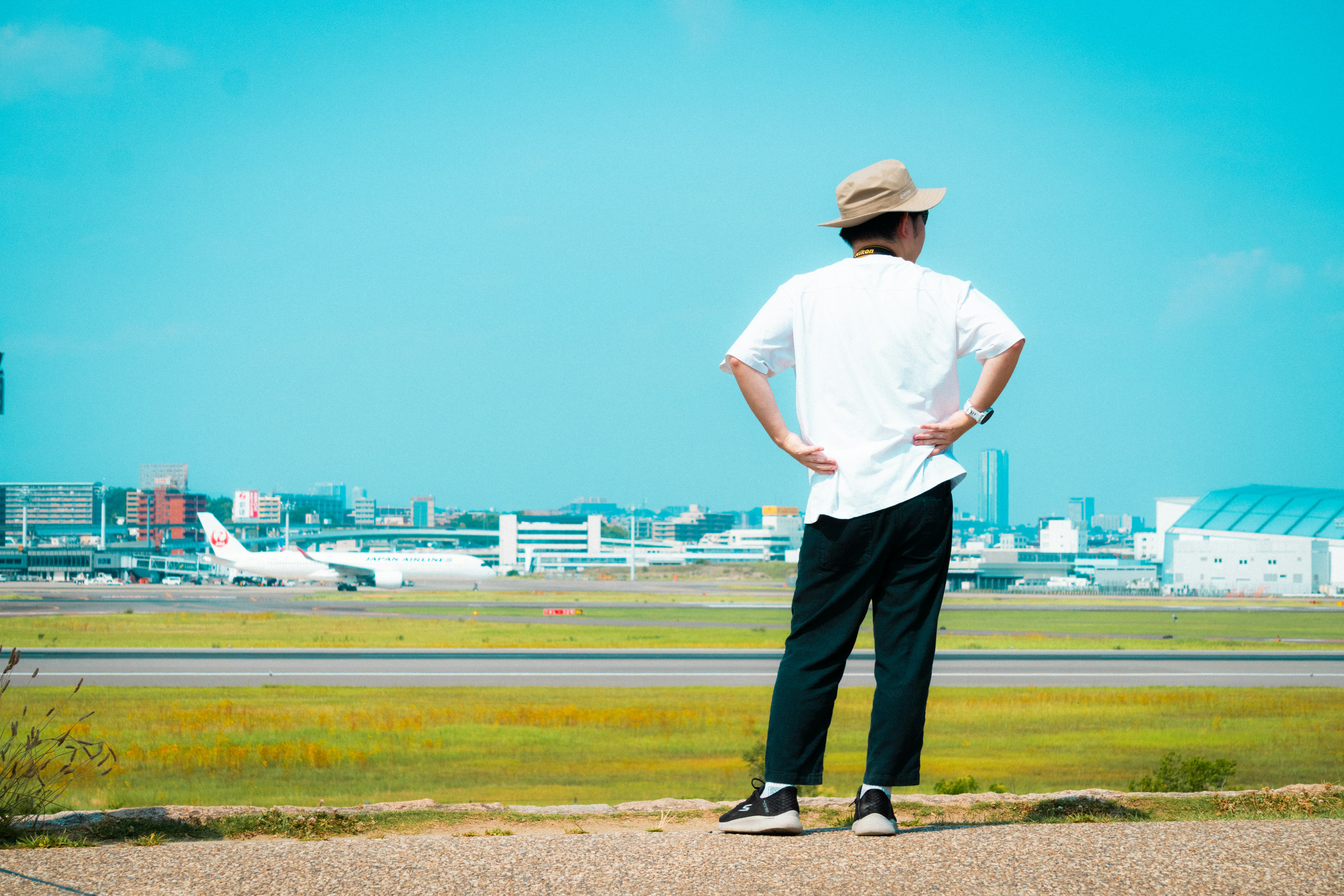 Person in a wide-brimmed hat gazing over an airport runway, with planes and city skyline in the background.